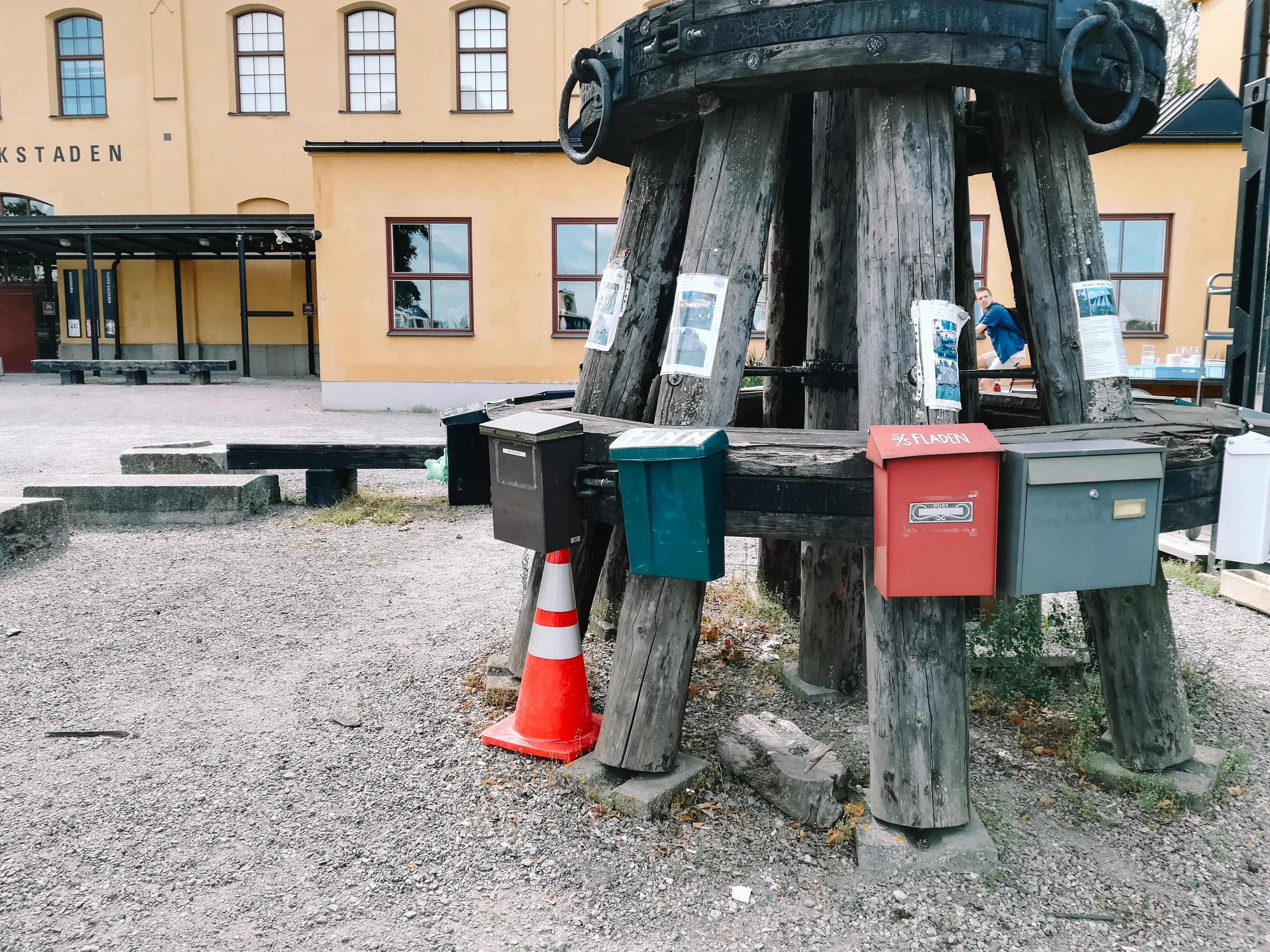 A rustic postal station featuring colorful mailboxes attached to a wooden structure, set against a backdrop of a historic building.