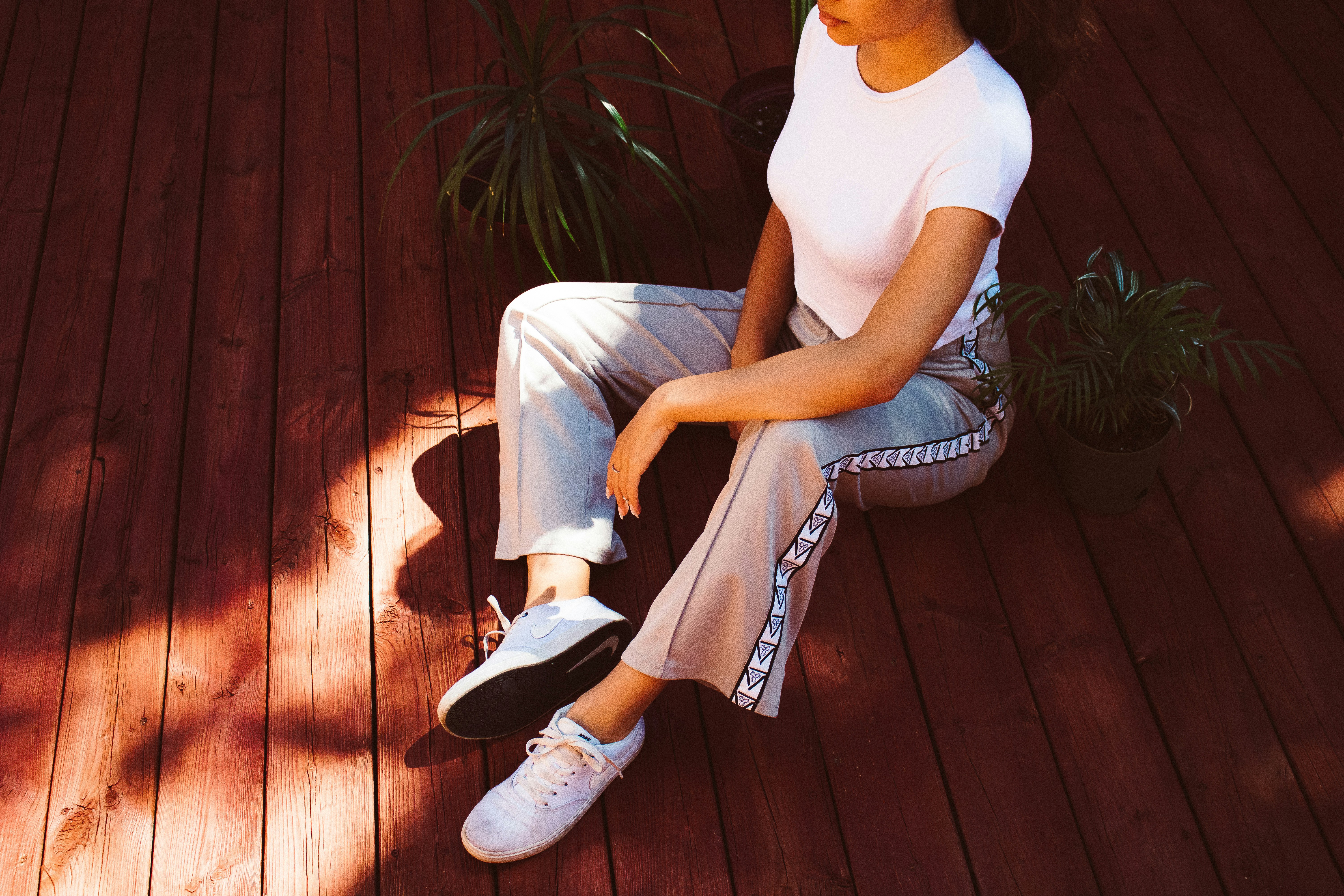A young woman sits casually on wooden flooring, surrounded by potted plants, with soft sunlight casting shadows around her.