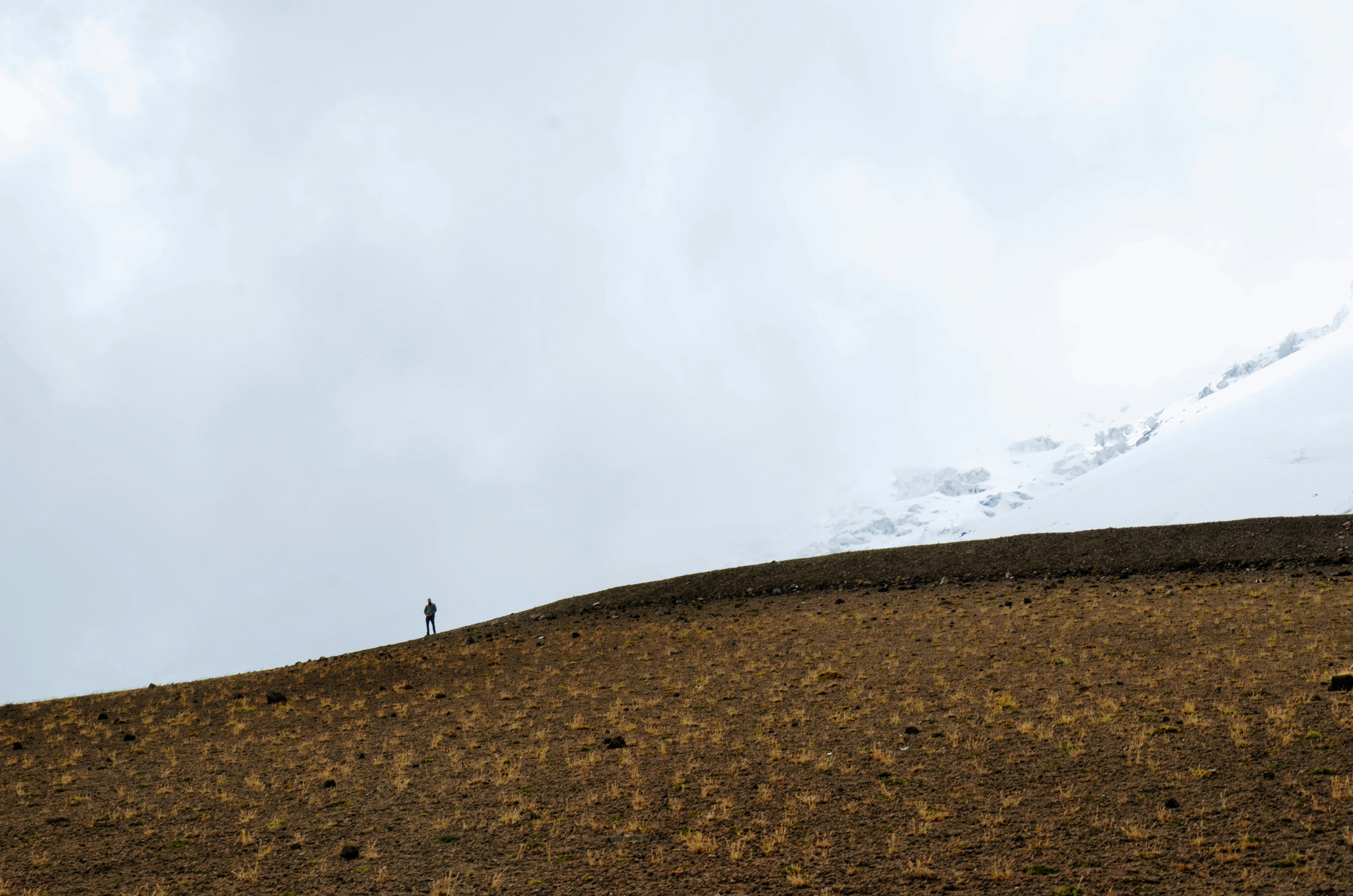 This striking image captures a lone hiker standing on a vast, barren mountain slope, emphasizing the scale and solitude of the landscape. The composition highlights the contrast between the earthy tones of the ground and the bright, overcast sky, creating a dramatic and contemplative atmosphere. The minimalist approach and the use of negative space make the image visually compelling, drawing the viewer's attention to the small figure amidst the expansive terrain.