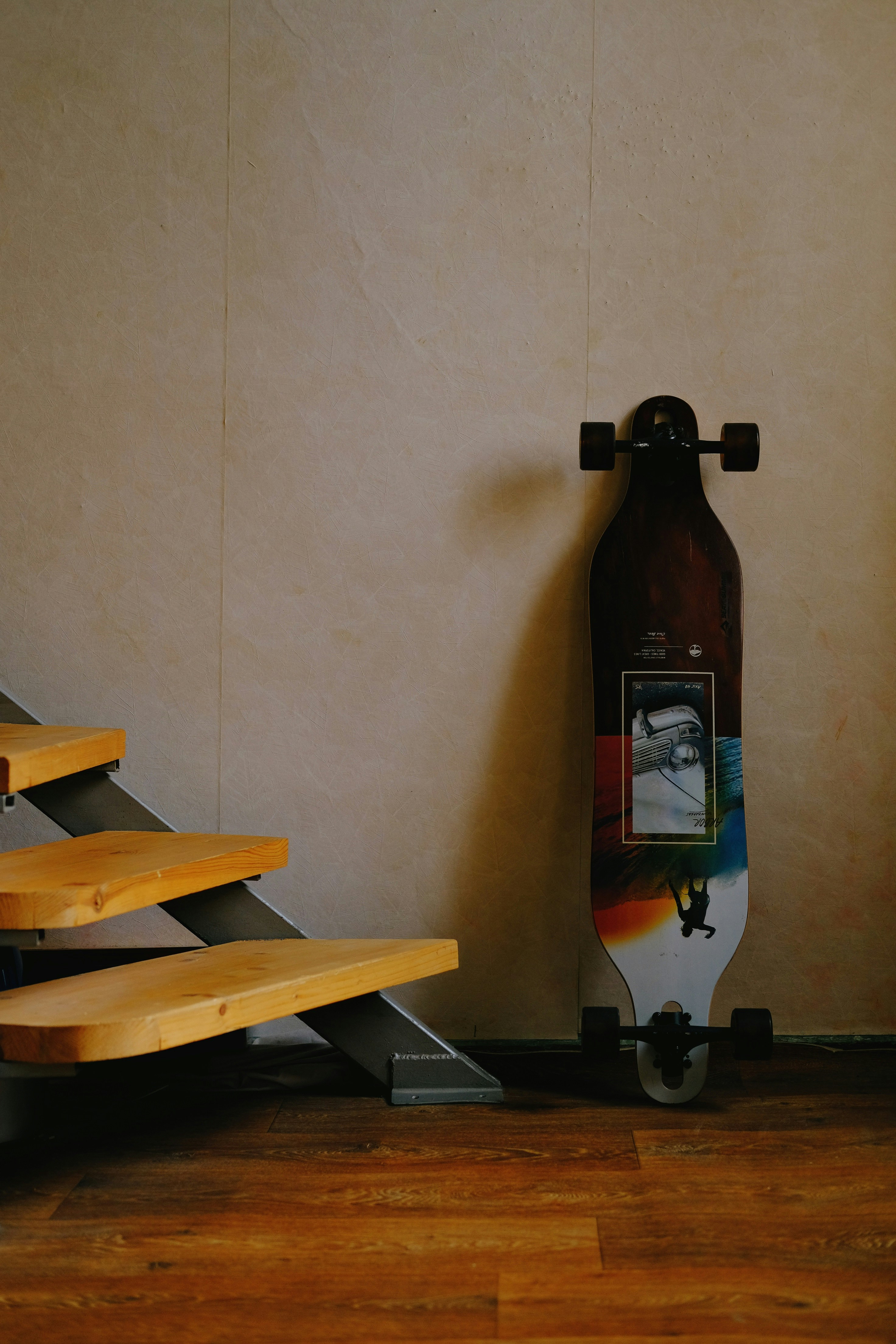 A longboard rests against a textured wall beside wooden stairs, showcasing its vibrant design and inviting a sense of calm in an urban setting.