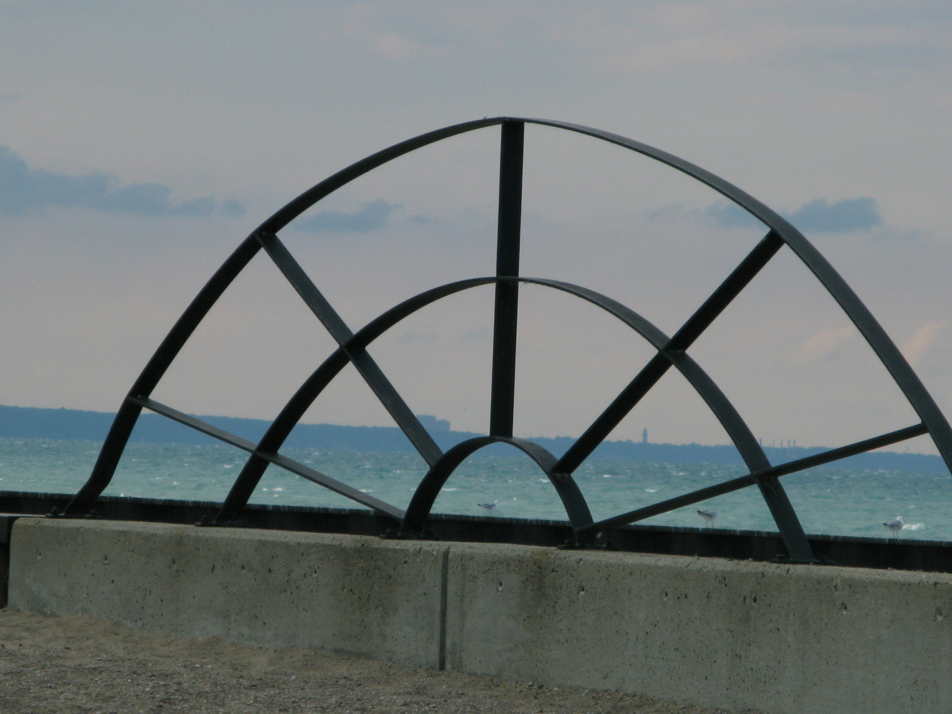 Concentric metal arches rise from a concrete seawall, framing the turquoise sea and distant horizon.