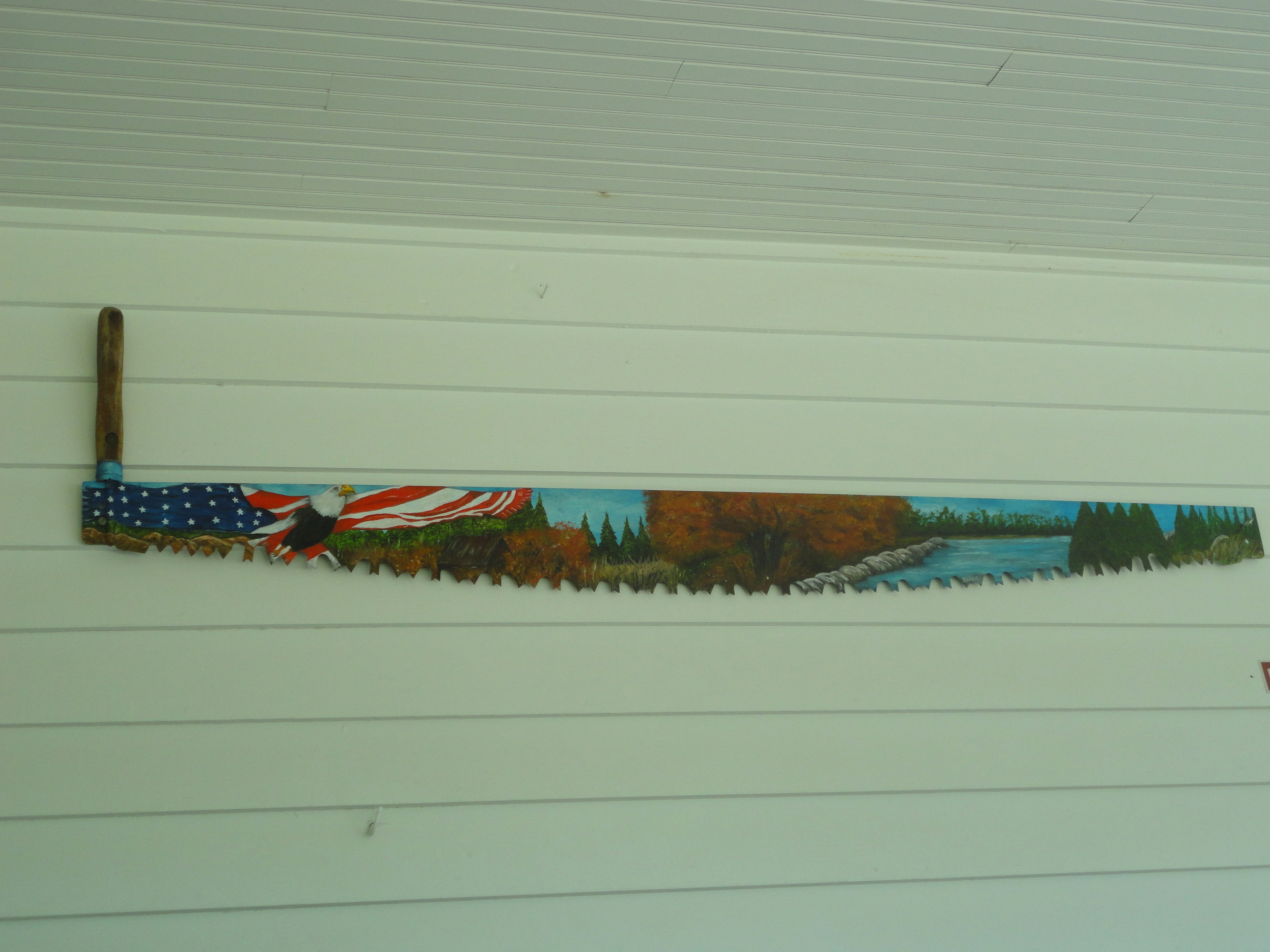 A creatively painted saw featuring a scenic landscape with trees, a river, and an American flag. The artwork blends functionality with artistic expression.