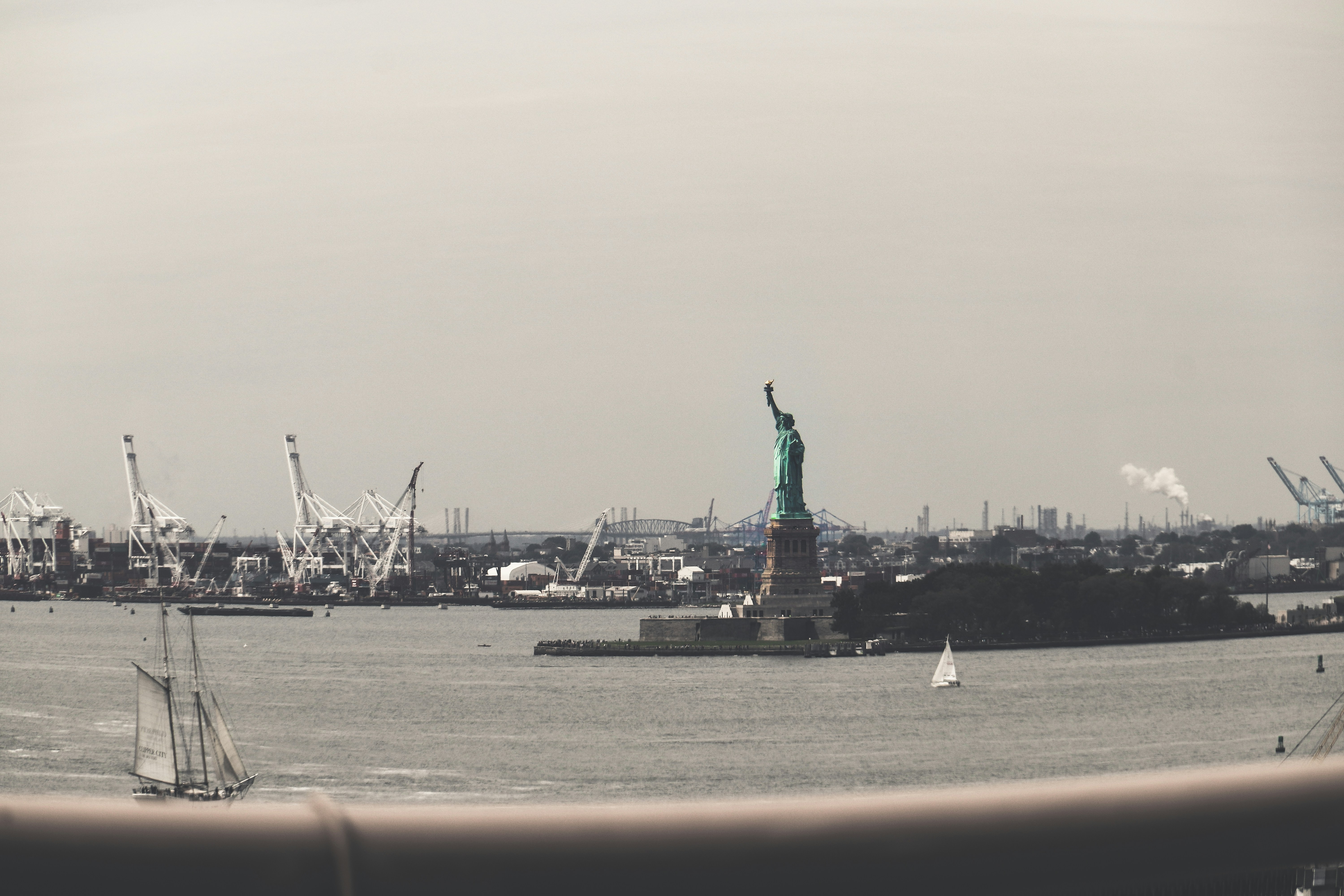 Statue of Liberty stands proudly in the harbor, surrounded by vessels and industrial cranes in the background.