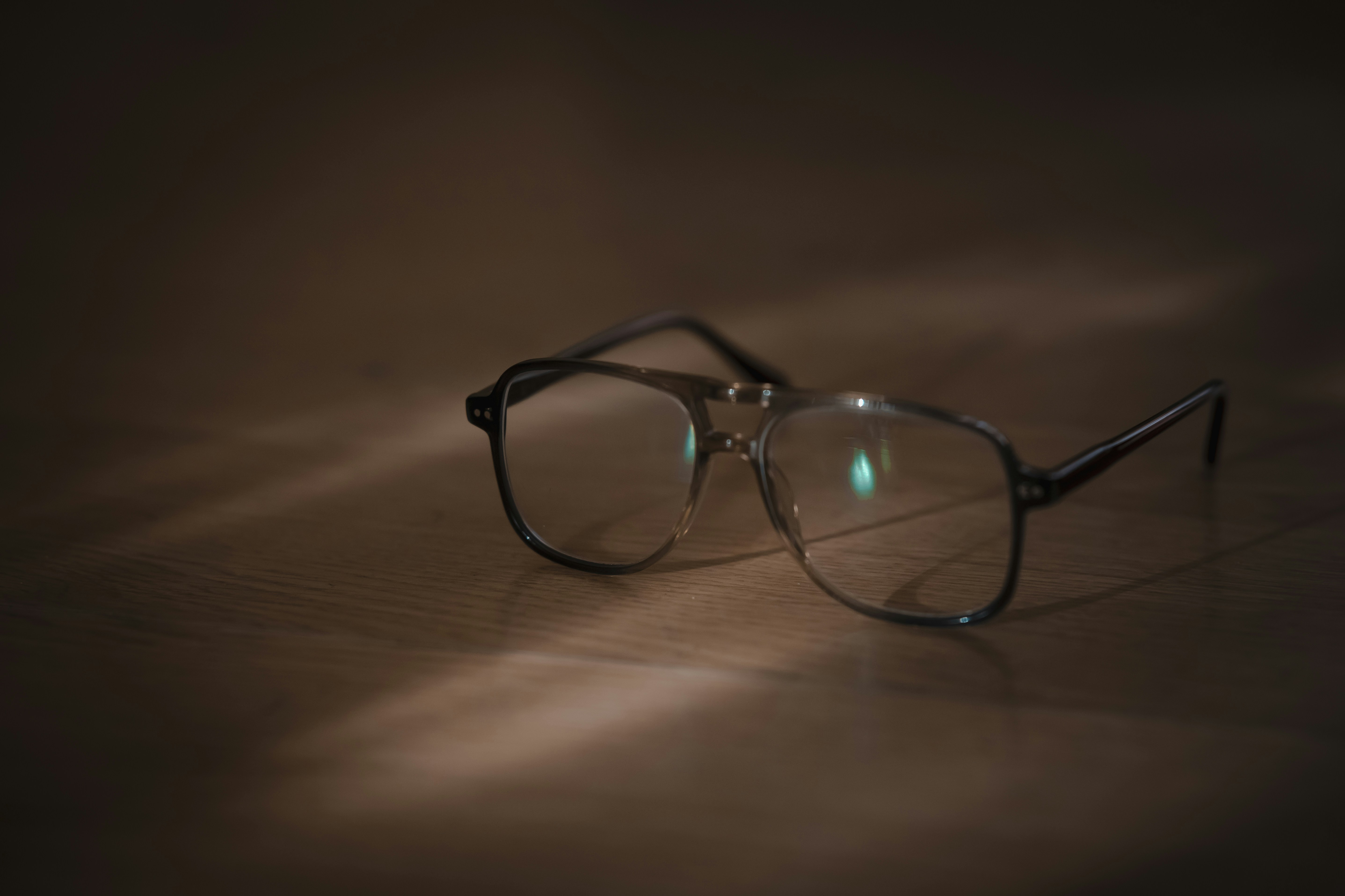A pair of eyeglasses rests on a wooden surface, illuminated by soft light that highlights their contours and reflections.