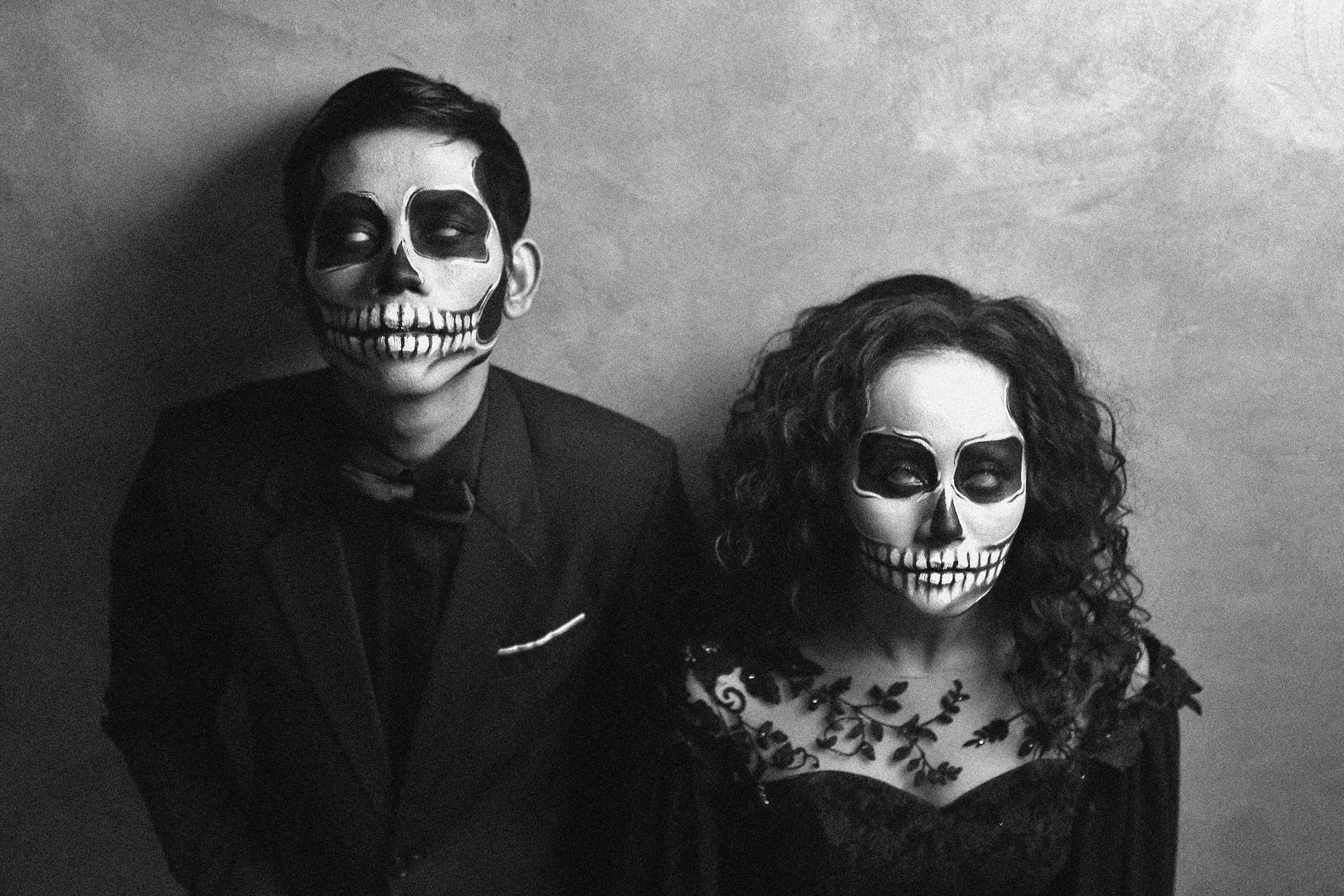 Man and woman with intricate skeleton makeup in formal attire against a textured backdrop.