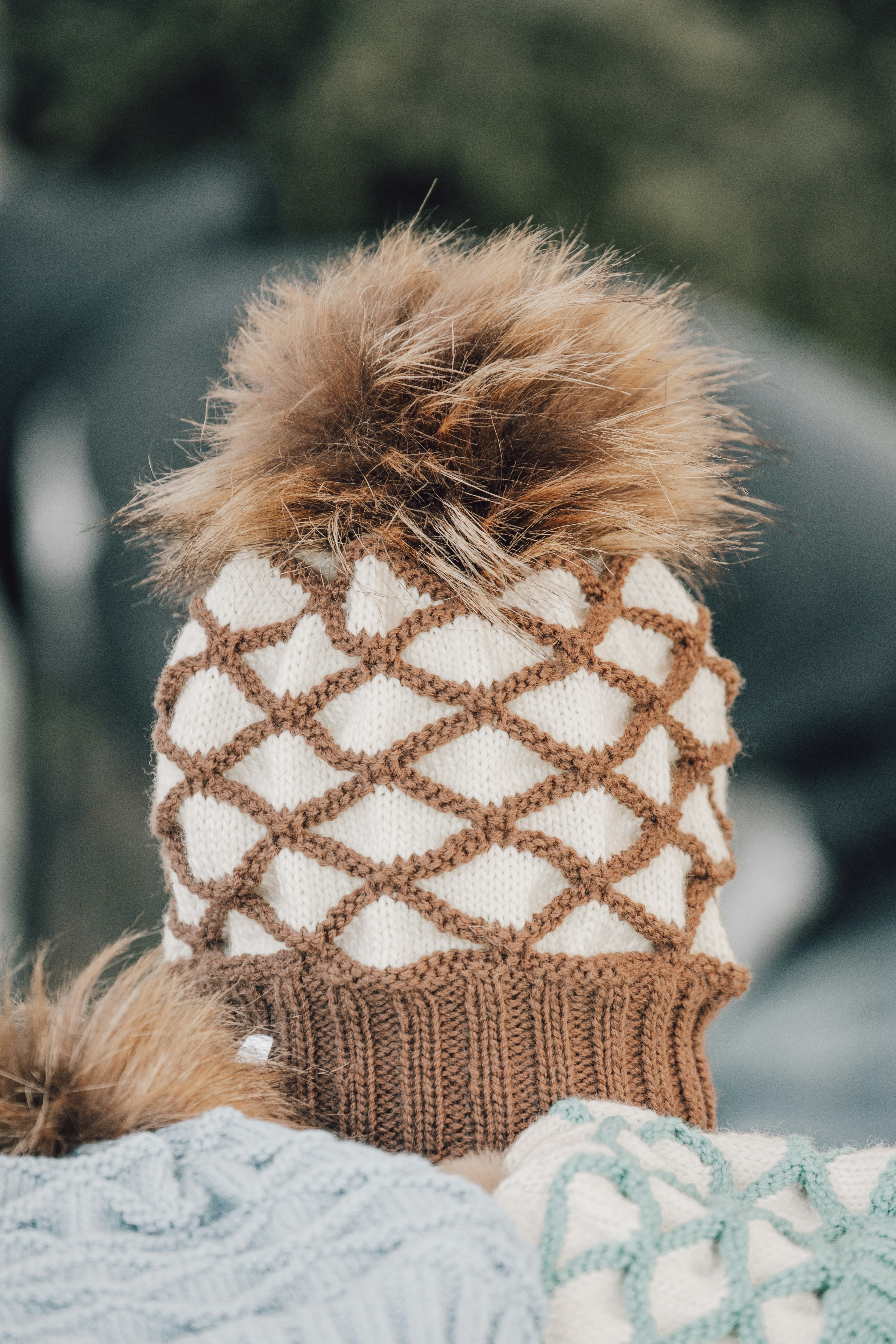 A knitted hat resembling a pineapple, featuring a fluffy pom-pom on top and intricate patterns. The background showcases additional knitwear in soft colors.