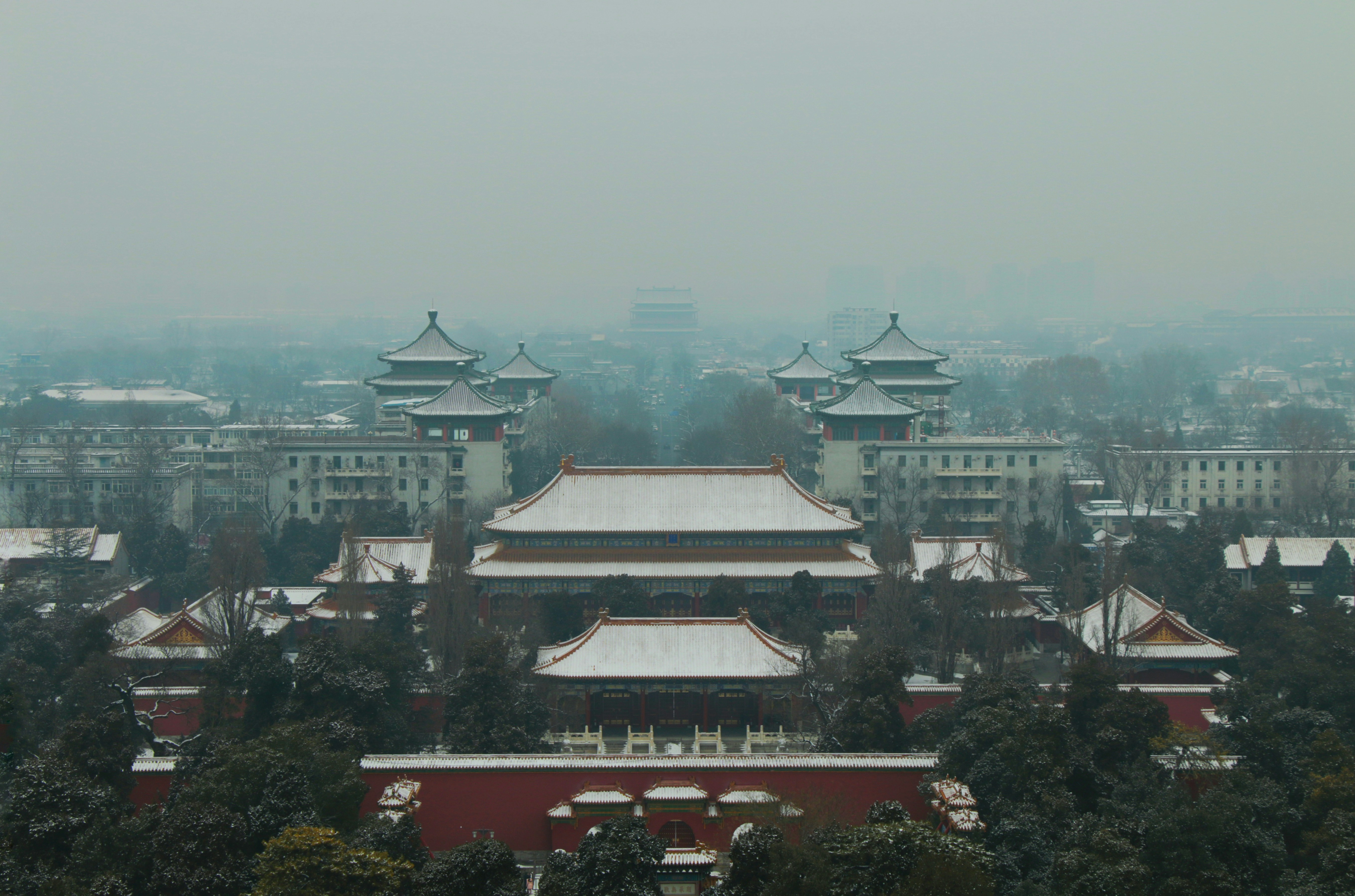 Aerial view of traditional Chinese architecture blanketed in snow, set against a hazy urban backdrop.