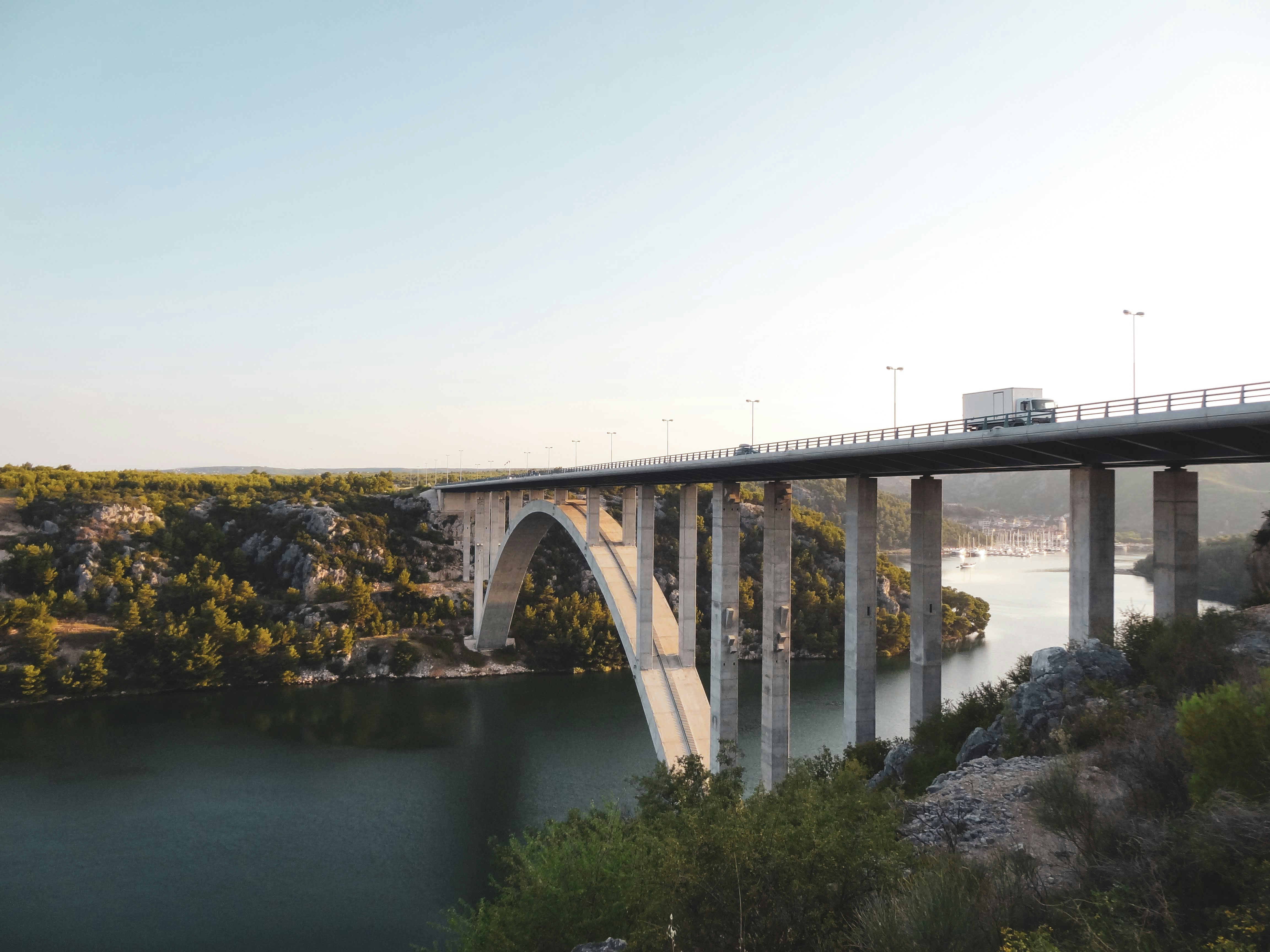 Long concrete arch bridge spans a calm river, with rocky cliffs and green scrub beneath a clear sky.