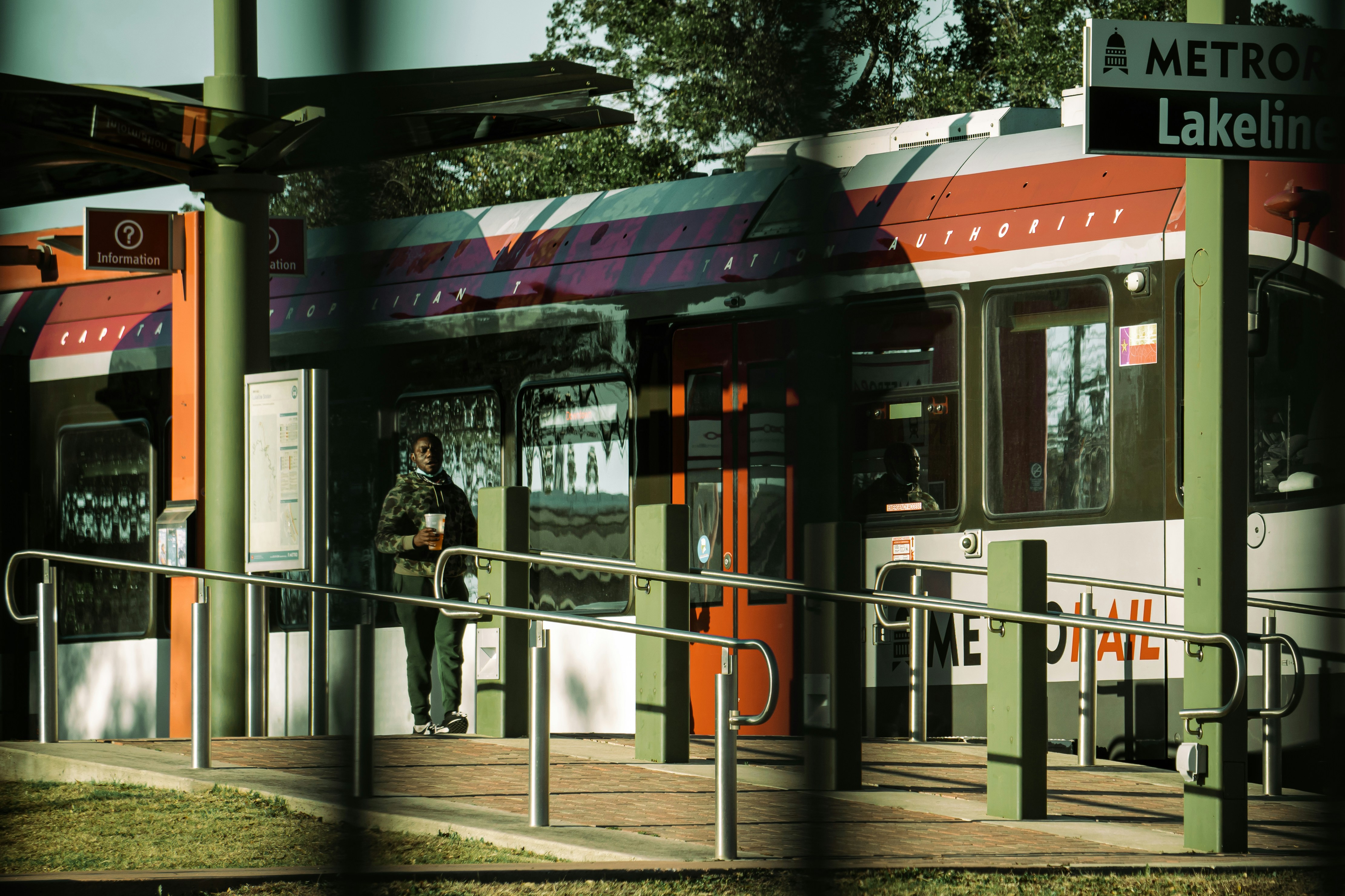 A lone figure stands at a transit station, awaiting the arrival of the metro train, with colorful structures framing the scene.