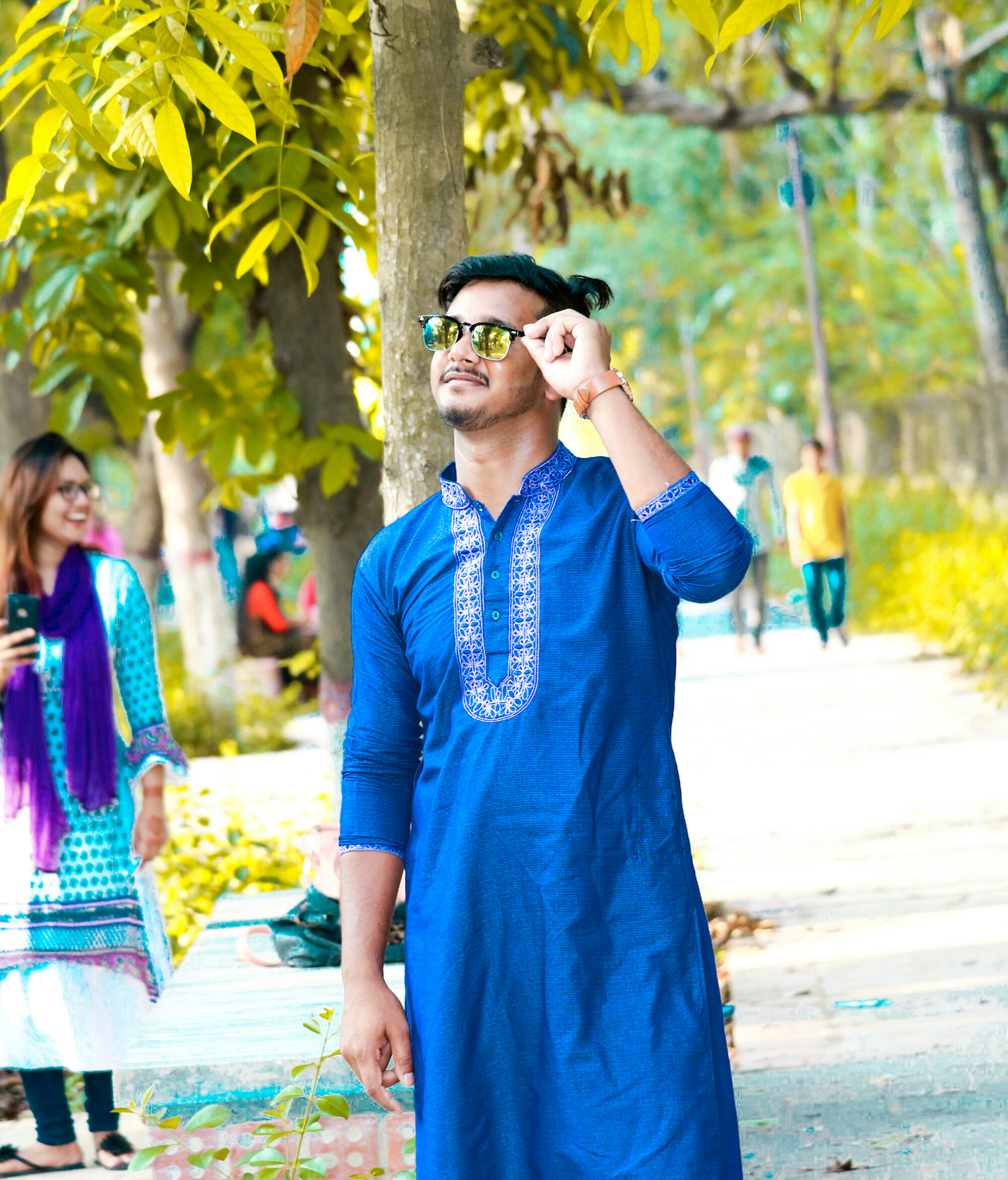 Man in a blue kurta adjusting his sunglasses, surrounded by lush greenery and friends enjoying a sunny day in the park.