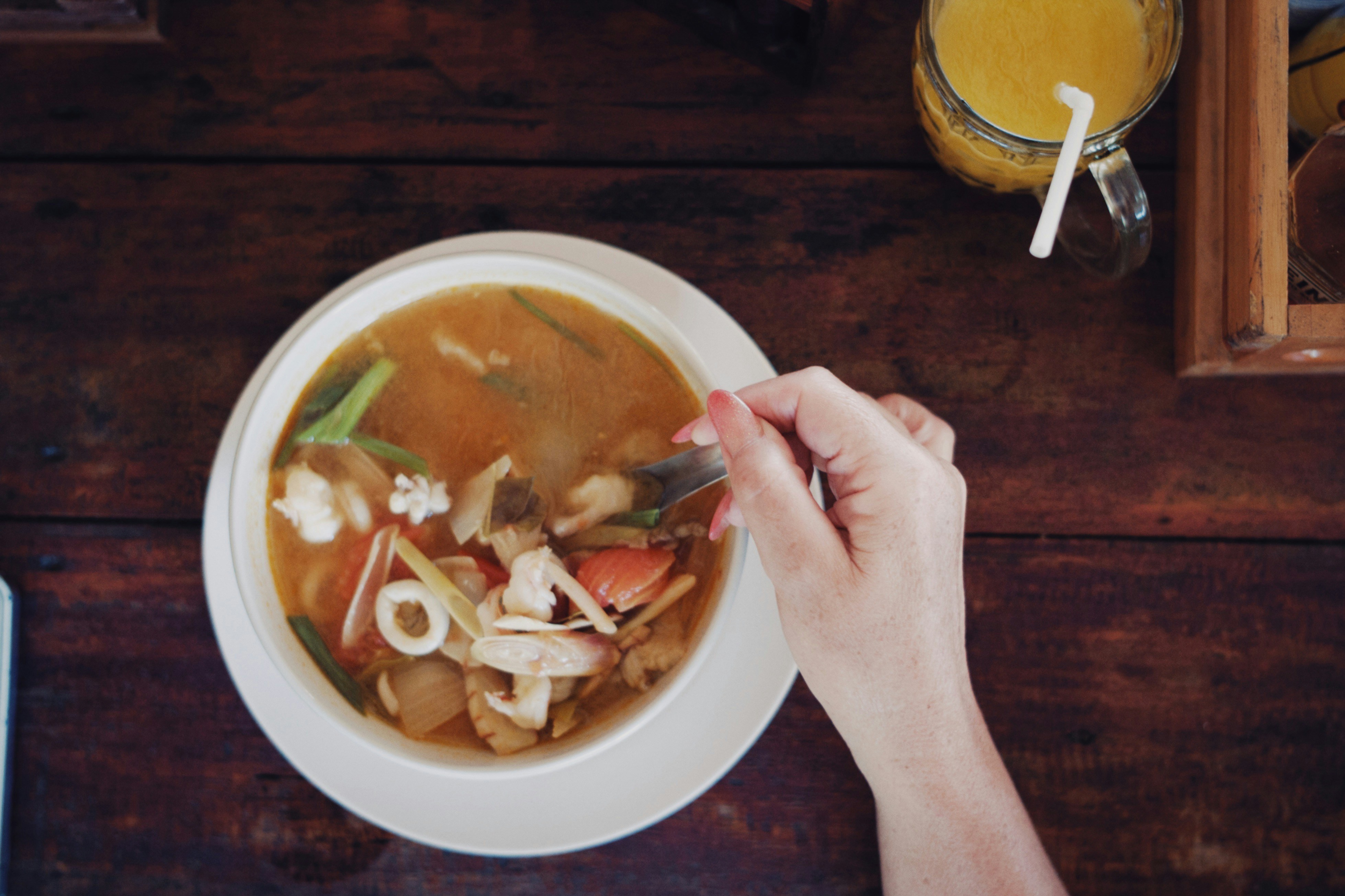 A hand delicately lifts a piece of seafood from a bowl of aromatic soup, showcasing vibrant ingredients on a rustic wooden table. A refreshing drink sits nearby.