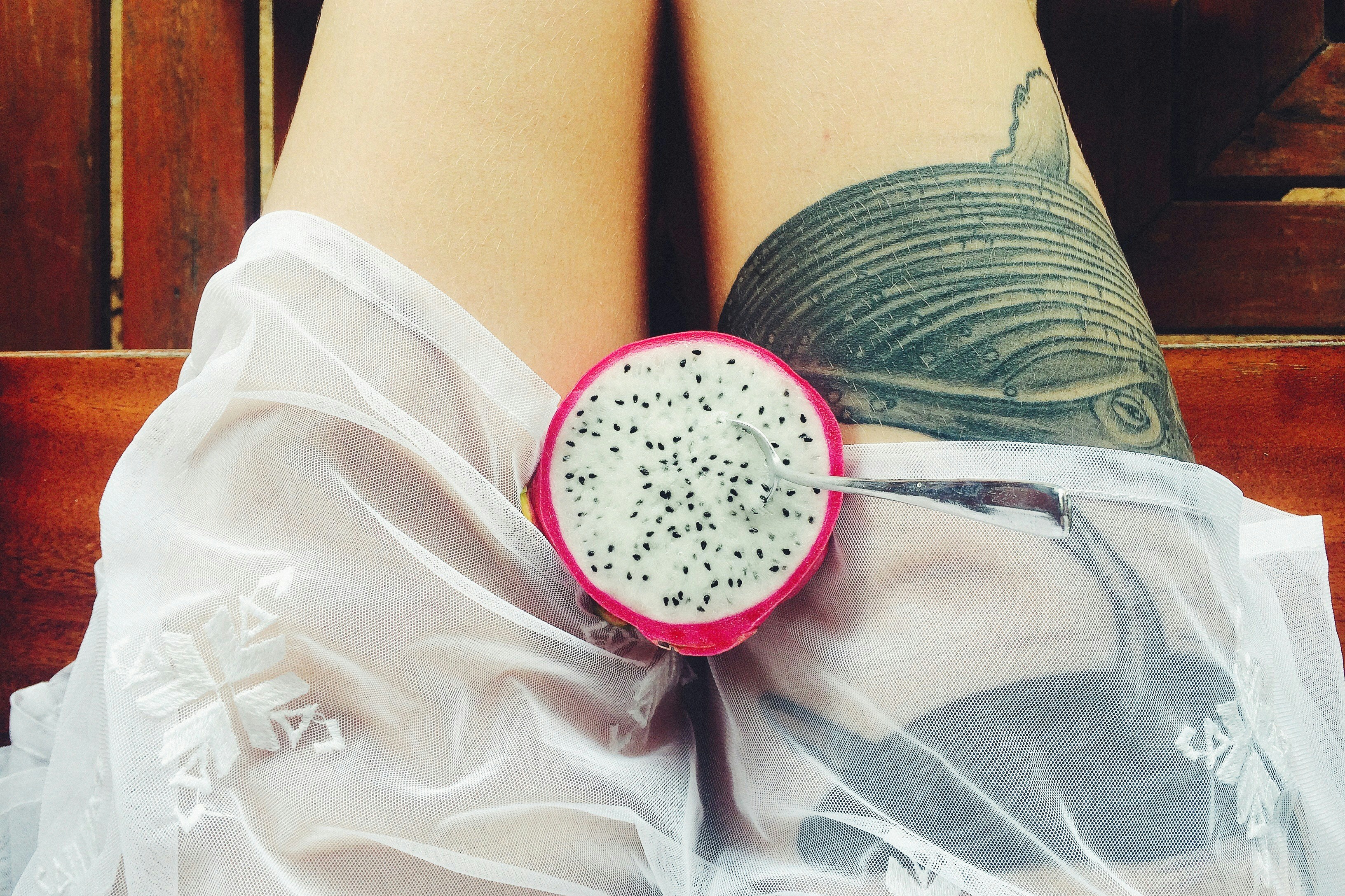 Close-up of a dragon fruit bowl resting on a person's thigh, adorned with a tattoo, against a wooden backdrop.