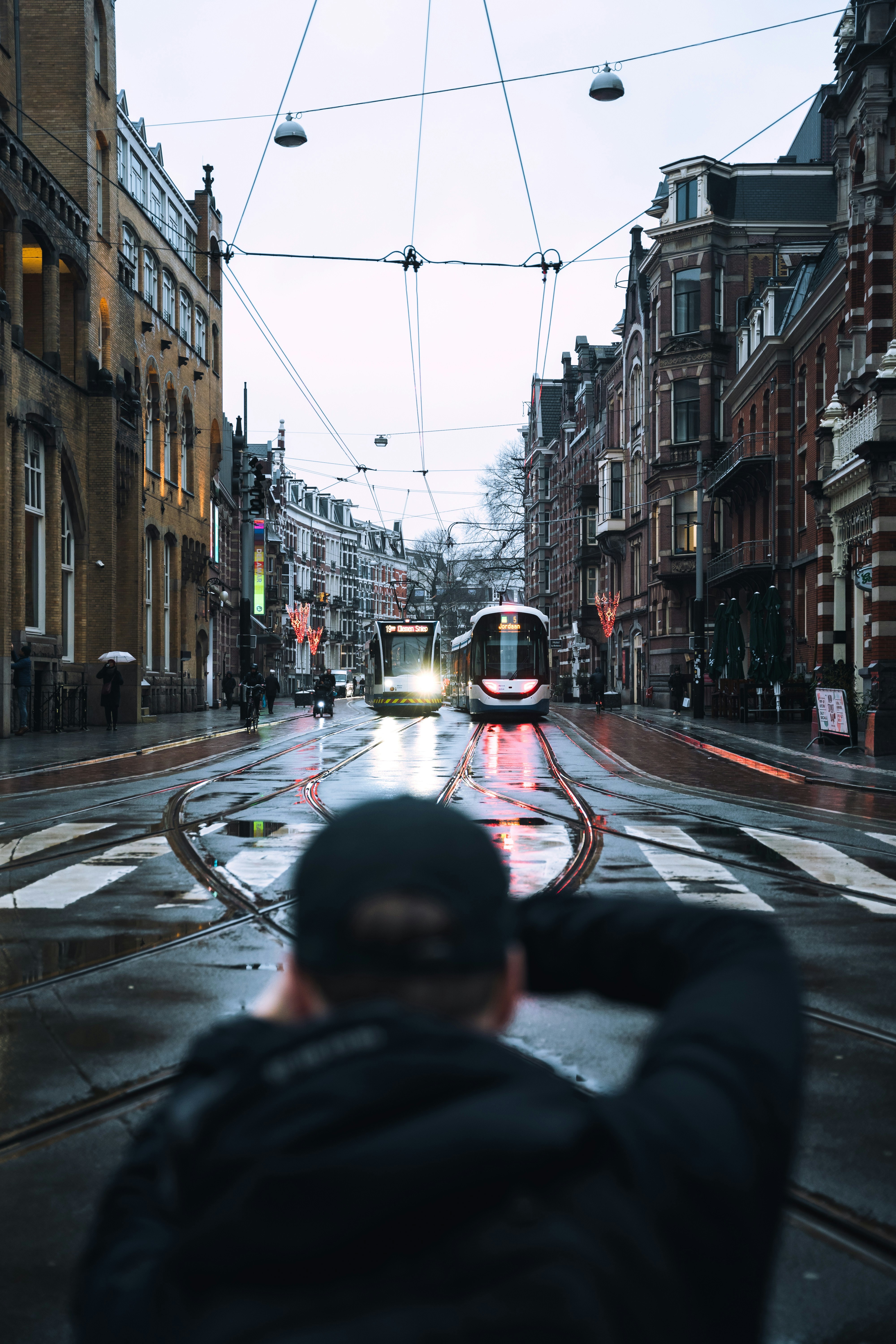 Two trams glide through a rain-soaked city street, reflecting vibrant lights against the wet pavement. A photographer captures the moment, adding a human element to the urban scene.