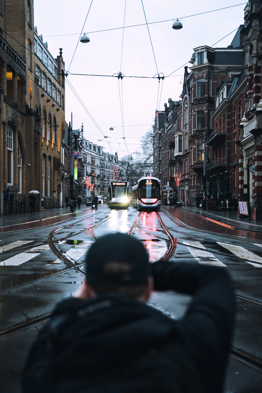 A blurred photographer in the foreground frames two trams on wet tracks between tall city buildings at dusk. Reflections and overhead wires lead the eye down the street.