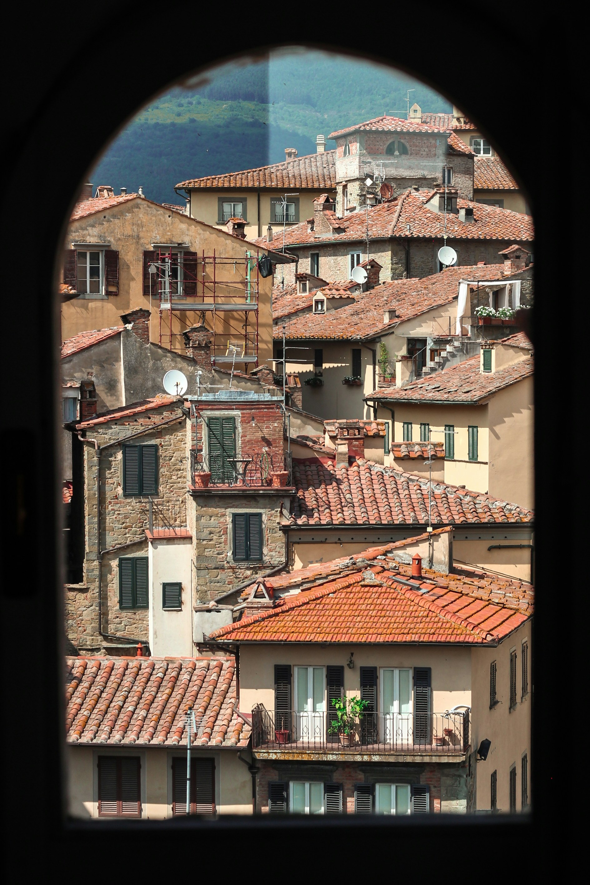 A view of rustic rooftops in a charming town, framed by an arched window. The scene showcases a mix of architectural styles and vibrant colors.