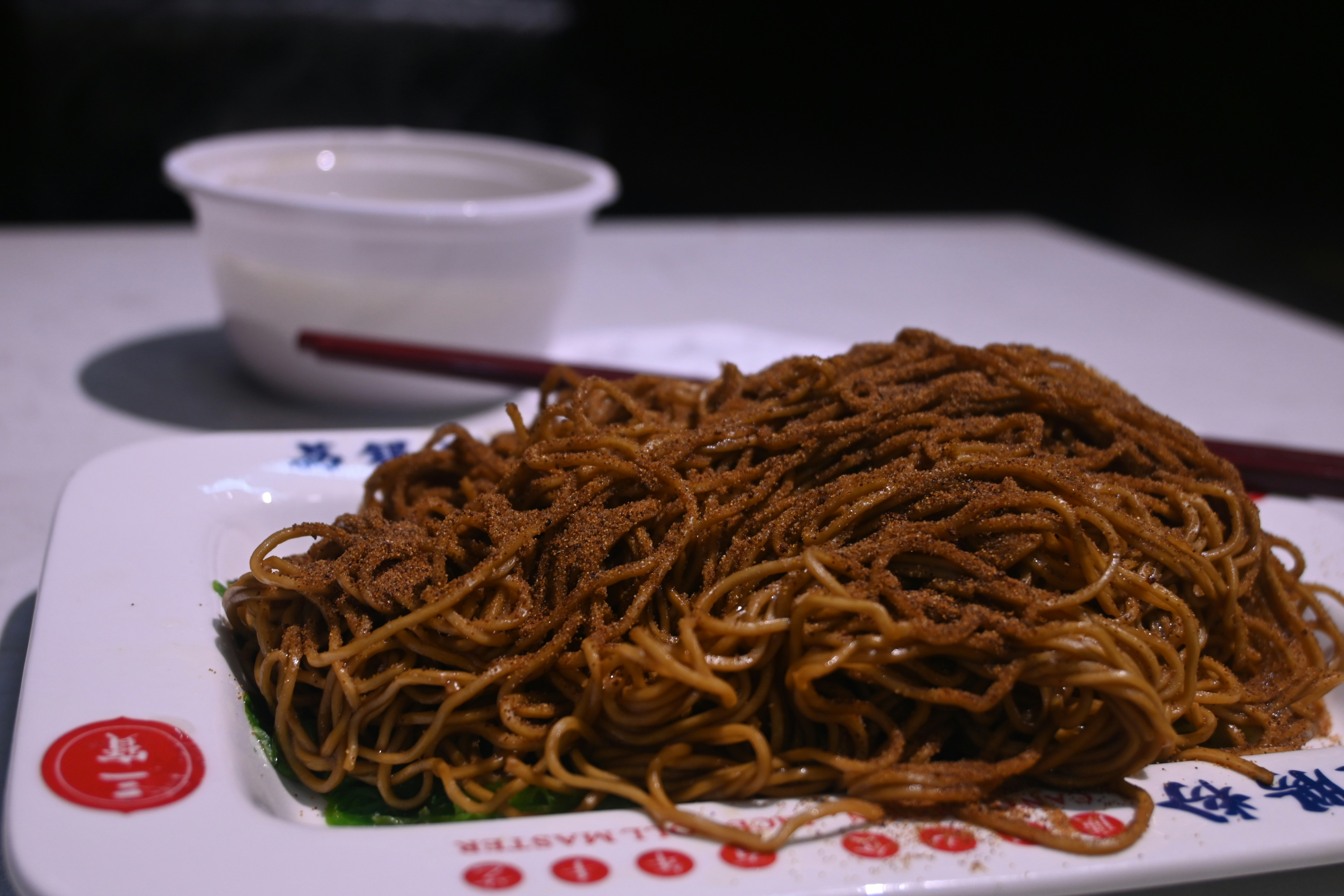 Plate of brown stir-fried noodles garnished with a side of sauce, set against a dimly lit background.