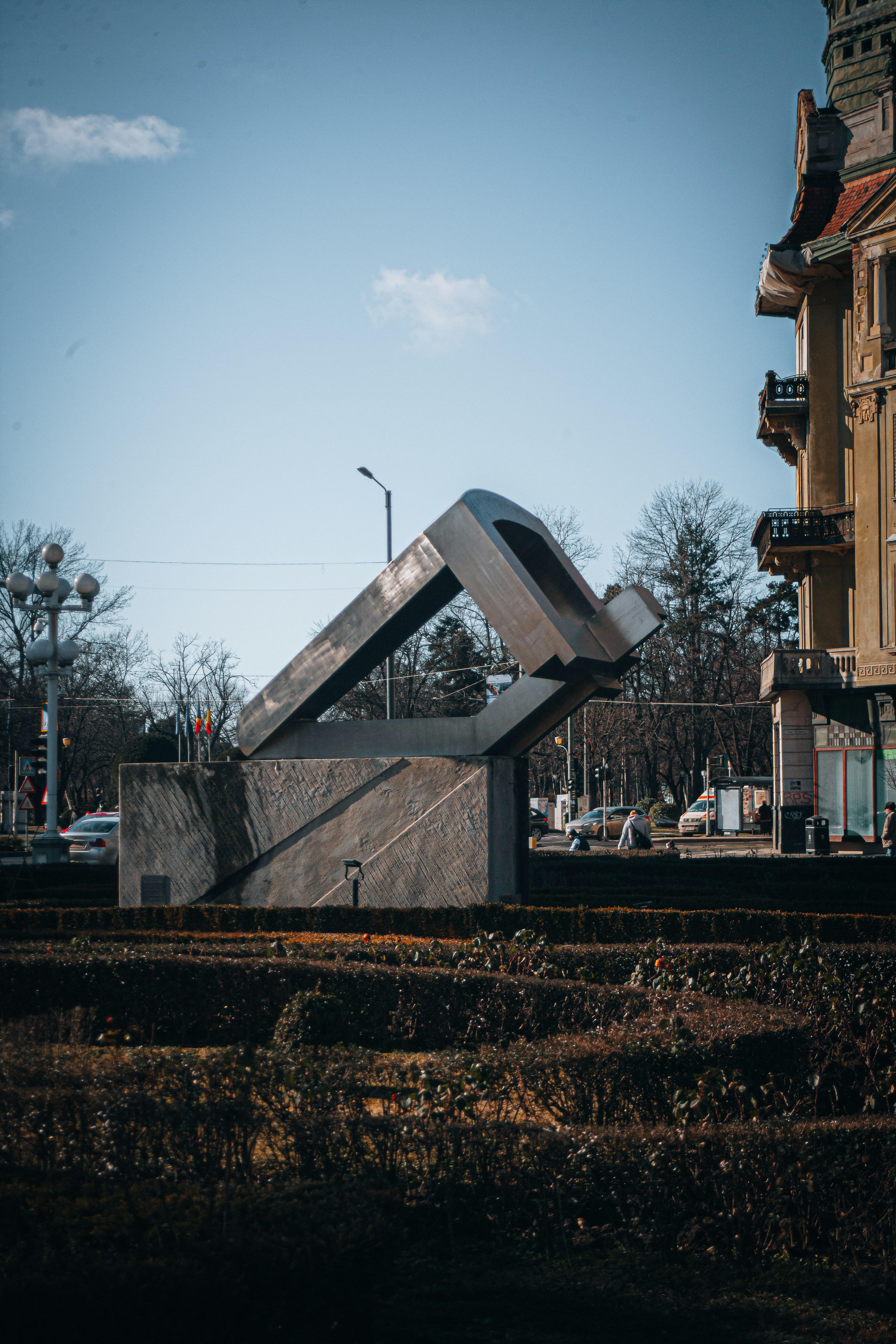 Geometric steel sculpture dominates a city square, framed by a classic building and bare hedges under clear daylight.