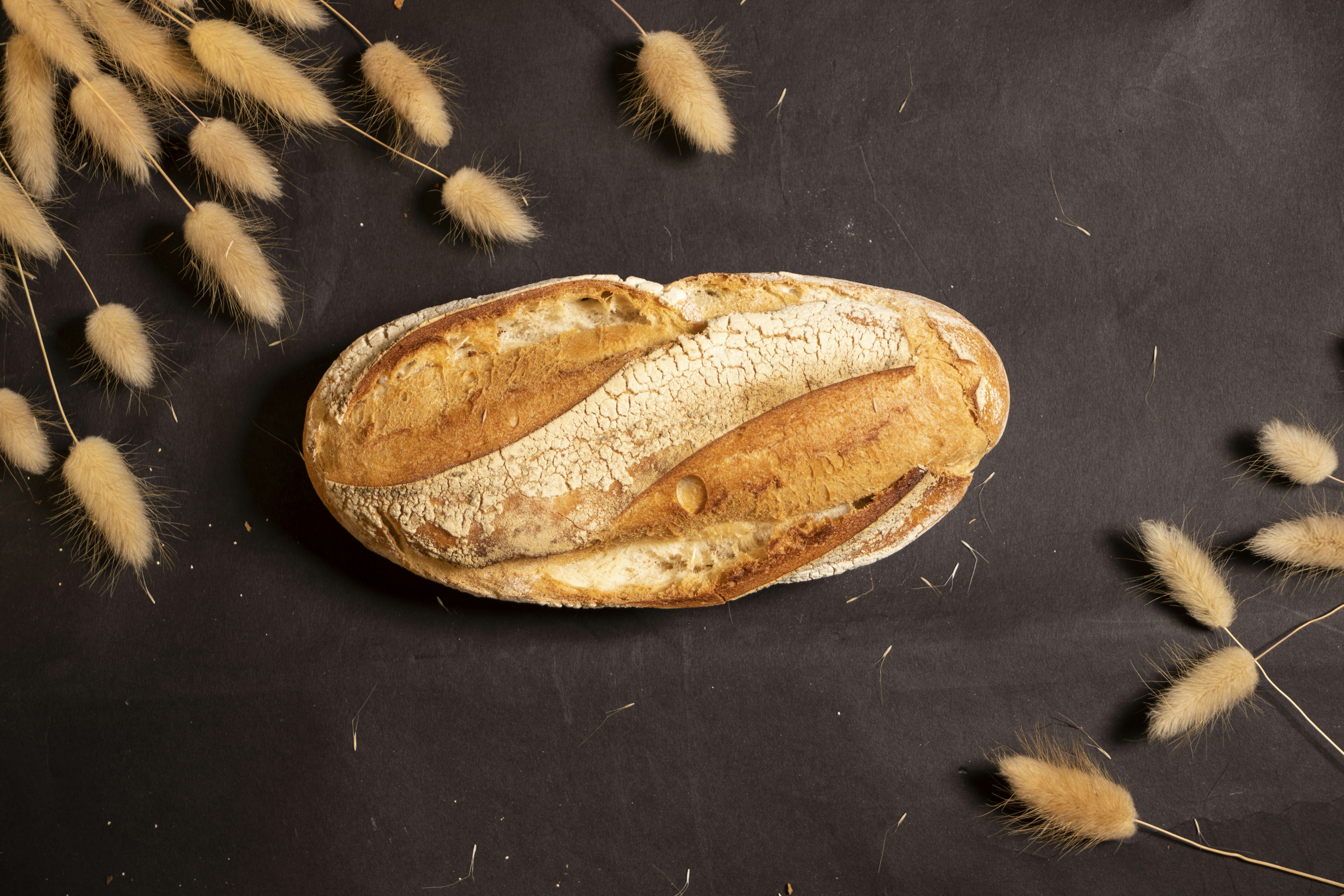 A rustic artisan bread loaf elegantly placed on a dark surface, surrounded by soft, feathery grasses.