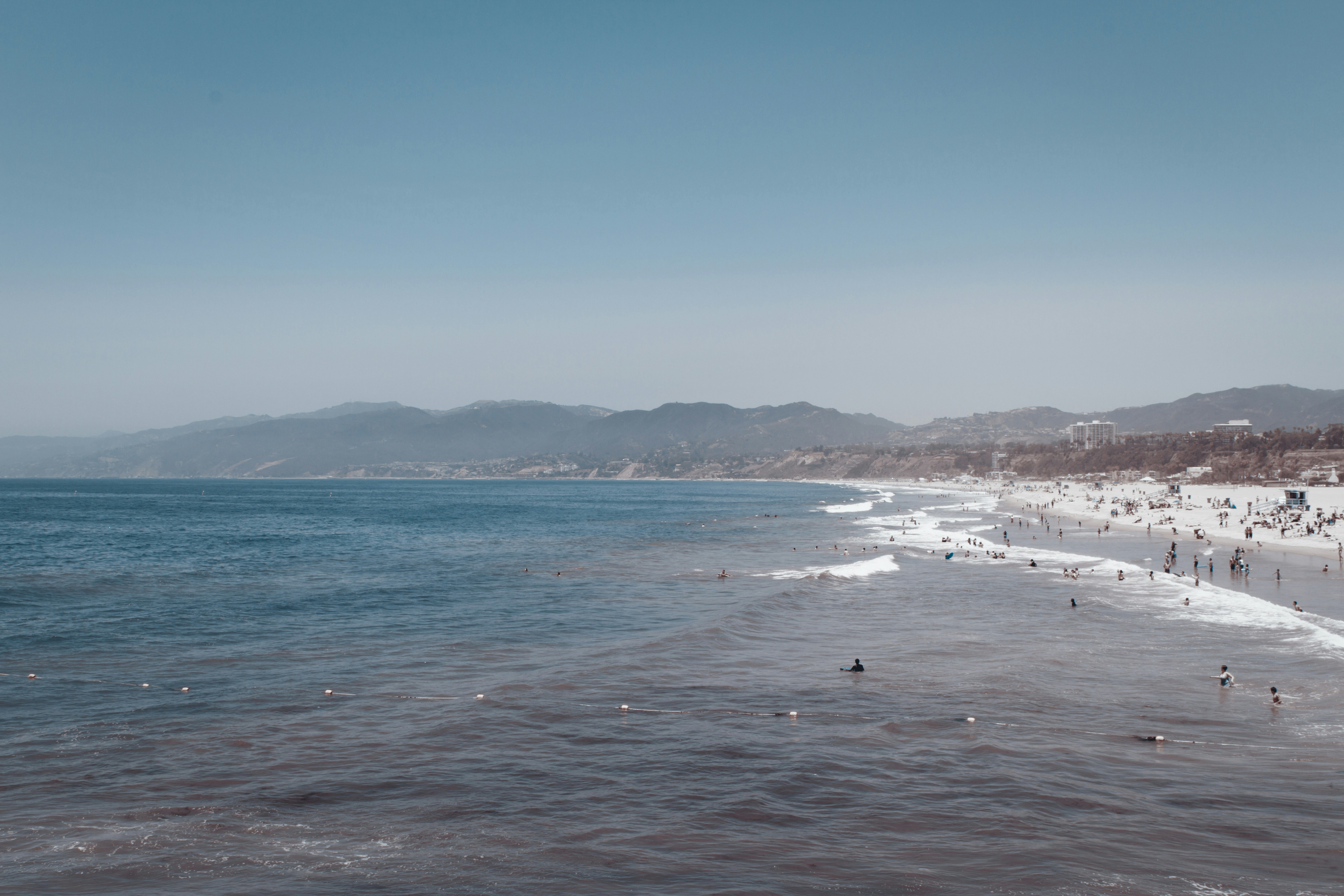Waves gently lap against the shore, where beachgoers enjoy a sunny day by the ocean. The distant mountains provide a serene backdrop to the bustling beach.