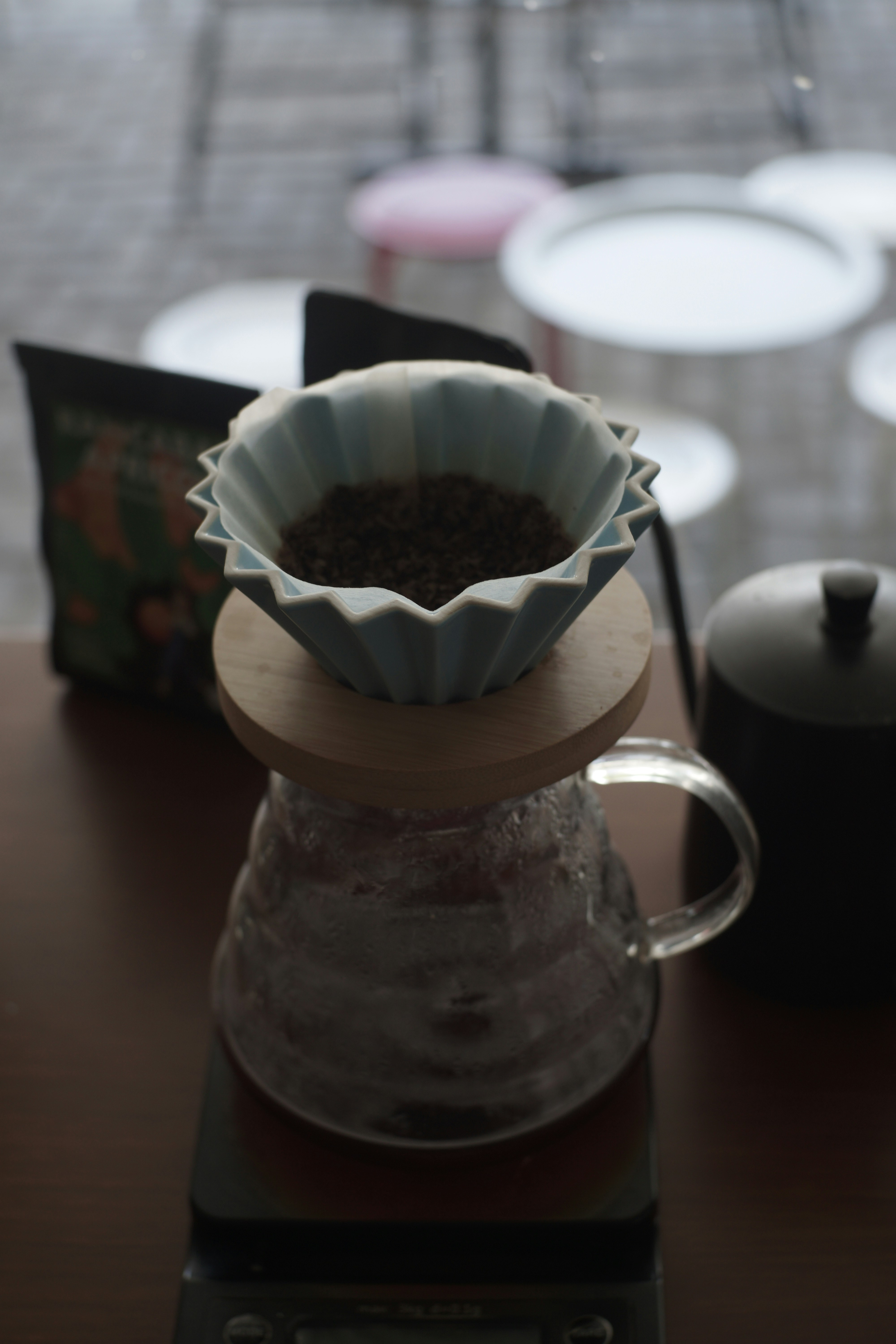 A coffee dripper filled with ground coffee sits atop a glass vessel, with blurred elements of a café in the background. The setup emphasizes the brewing process.