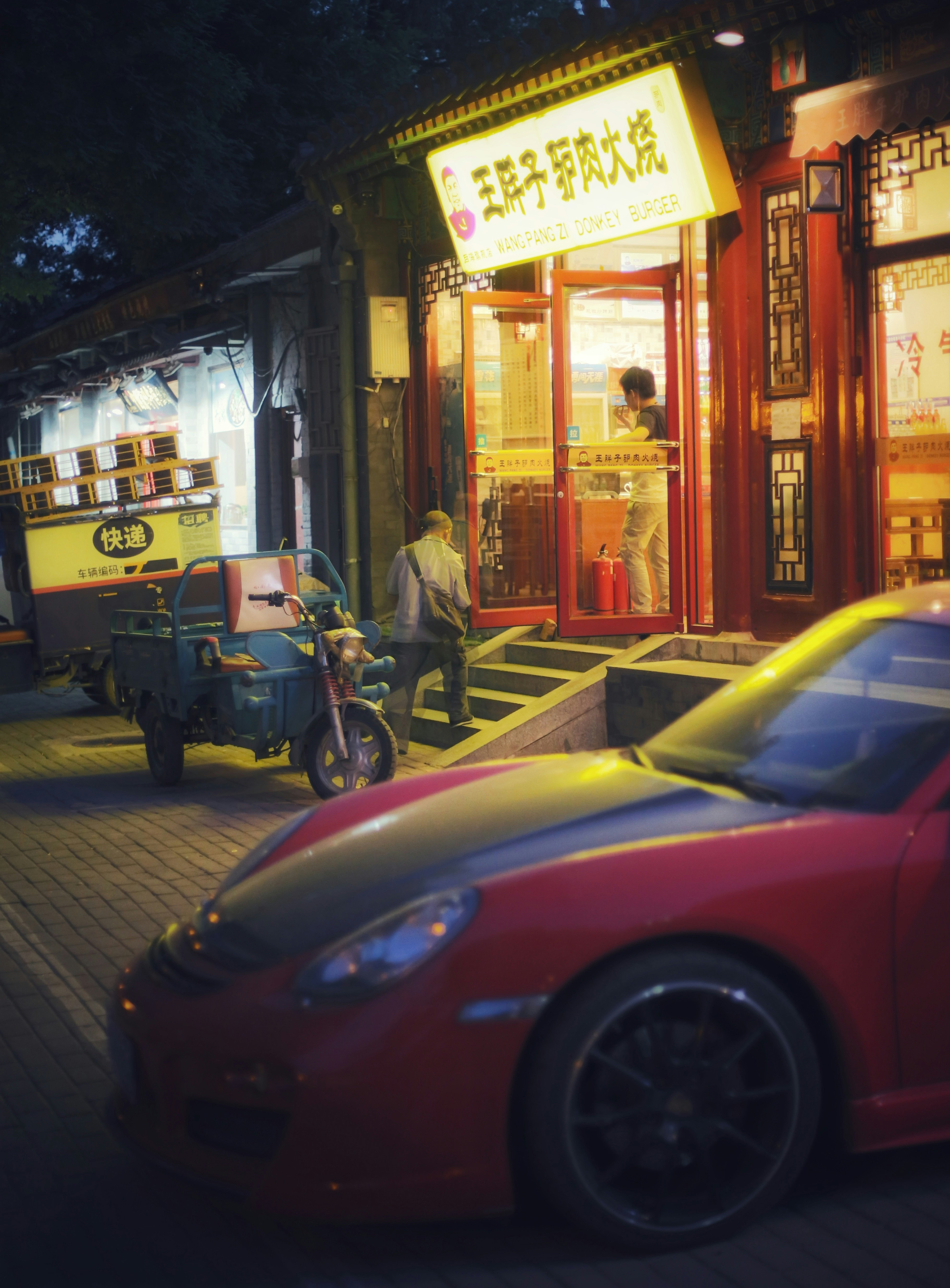 Crimson Car Meets Neon Threshold: Nightlife at a Lantern-Lit Shopfront