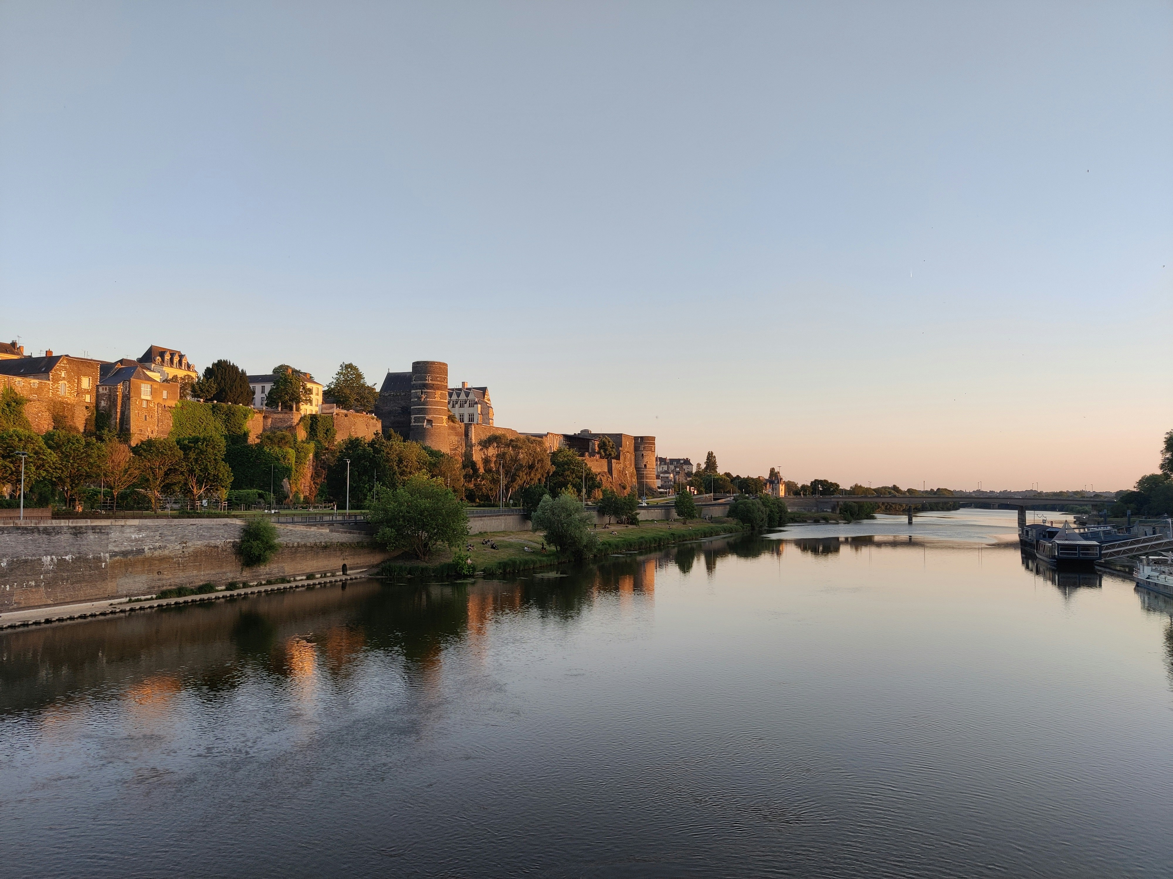 Historic buildings line a tranquil riverbank under a gradient evening sky, with a distant bridge in view.