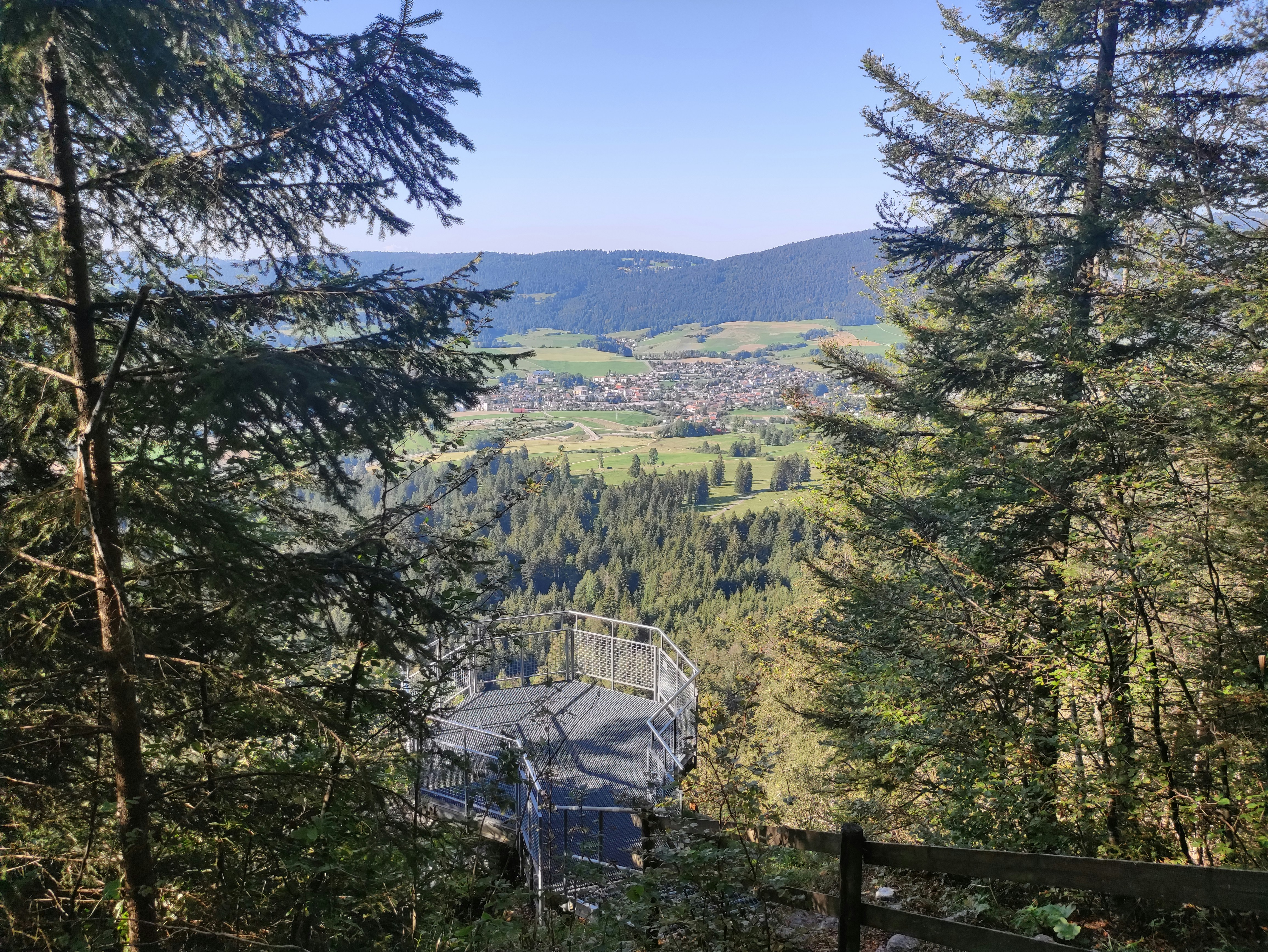 Circular viewing platform nestled among conifers overlooks a patchwork valley with a distant village and rolling hills.