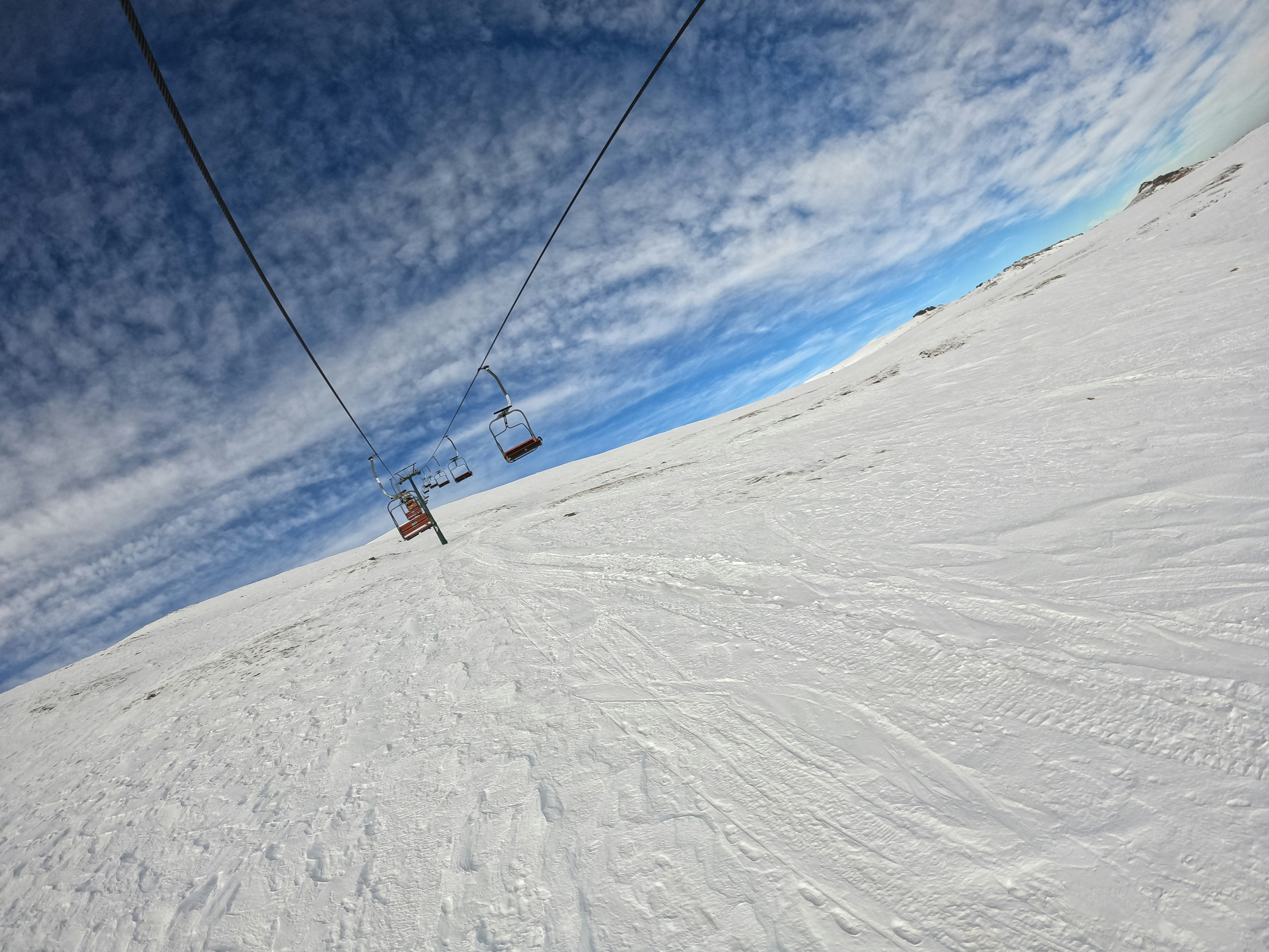 Powder-covered ski slope with a diagonal chairlift extending toward the horizon beneath a bright, partly cloudy sky. The wide-angle perspective emphasizes depth and movement along the lift line.