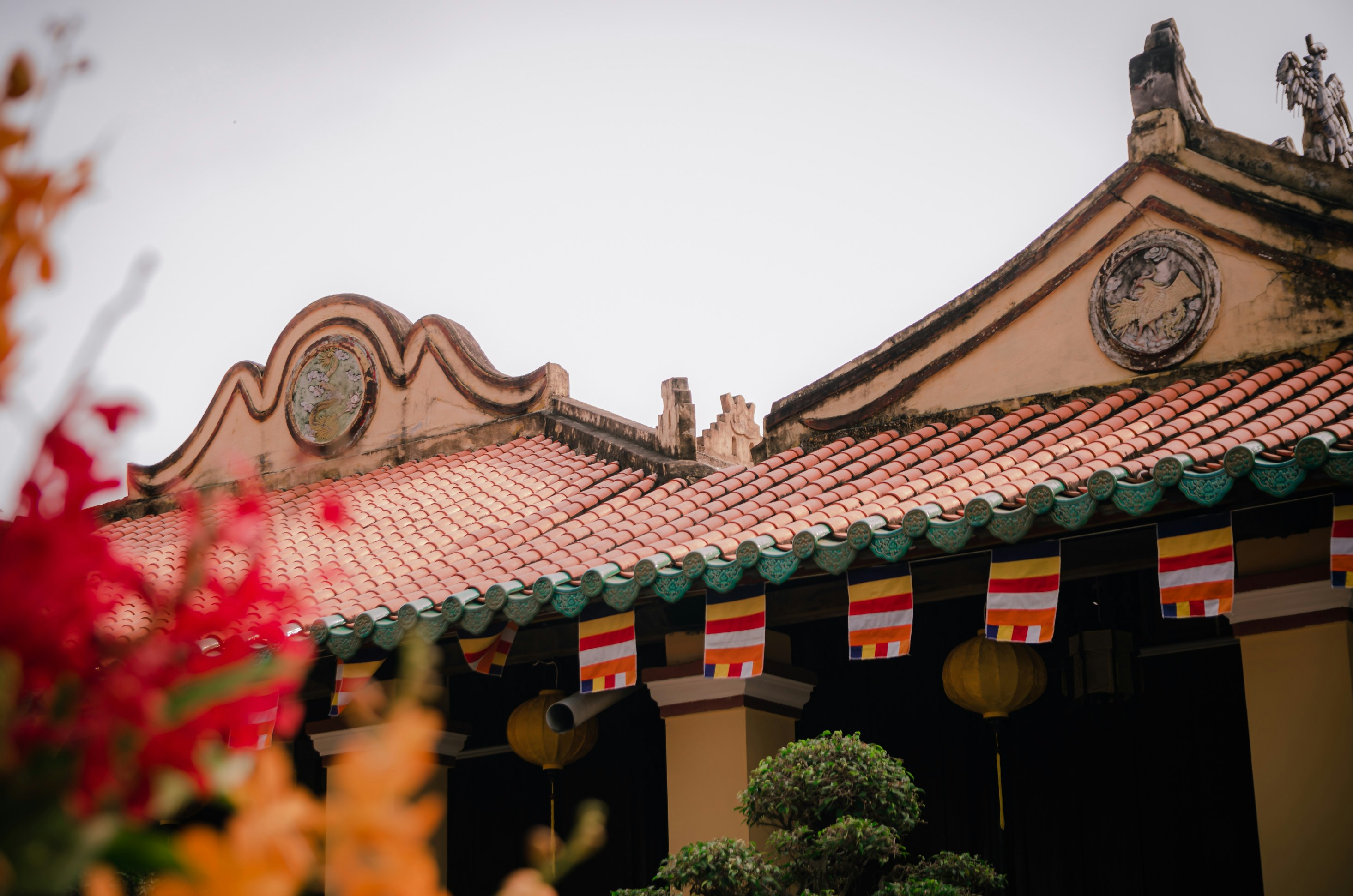 Traditional temple rooftops adorned with colorful banners and intricate carvings, framed by vibrant floral elements in the foreground.
