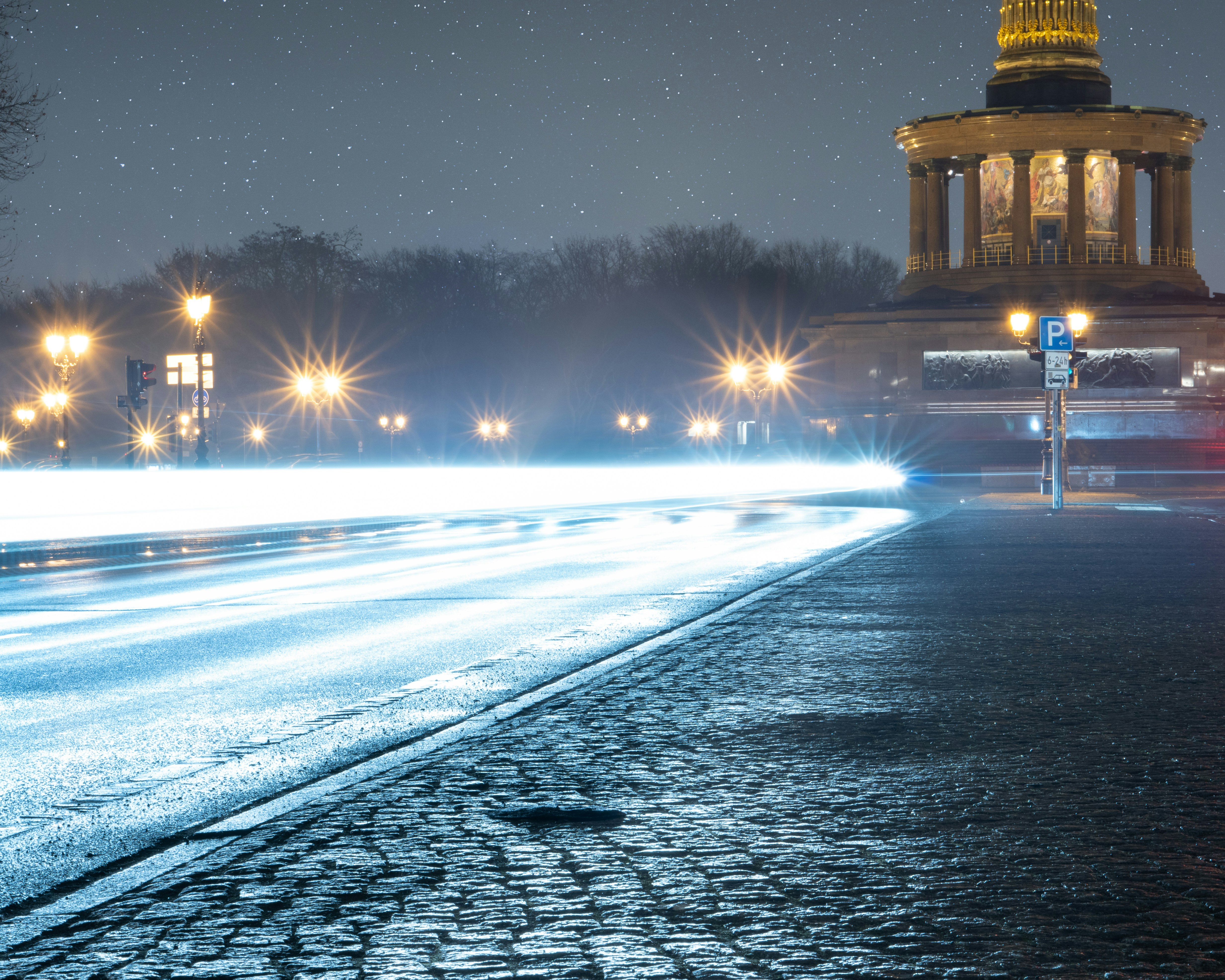 A vibrant night scene featuring light trails from passing vehicles alongside a historic monument, set against a starry sky.