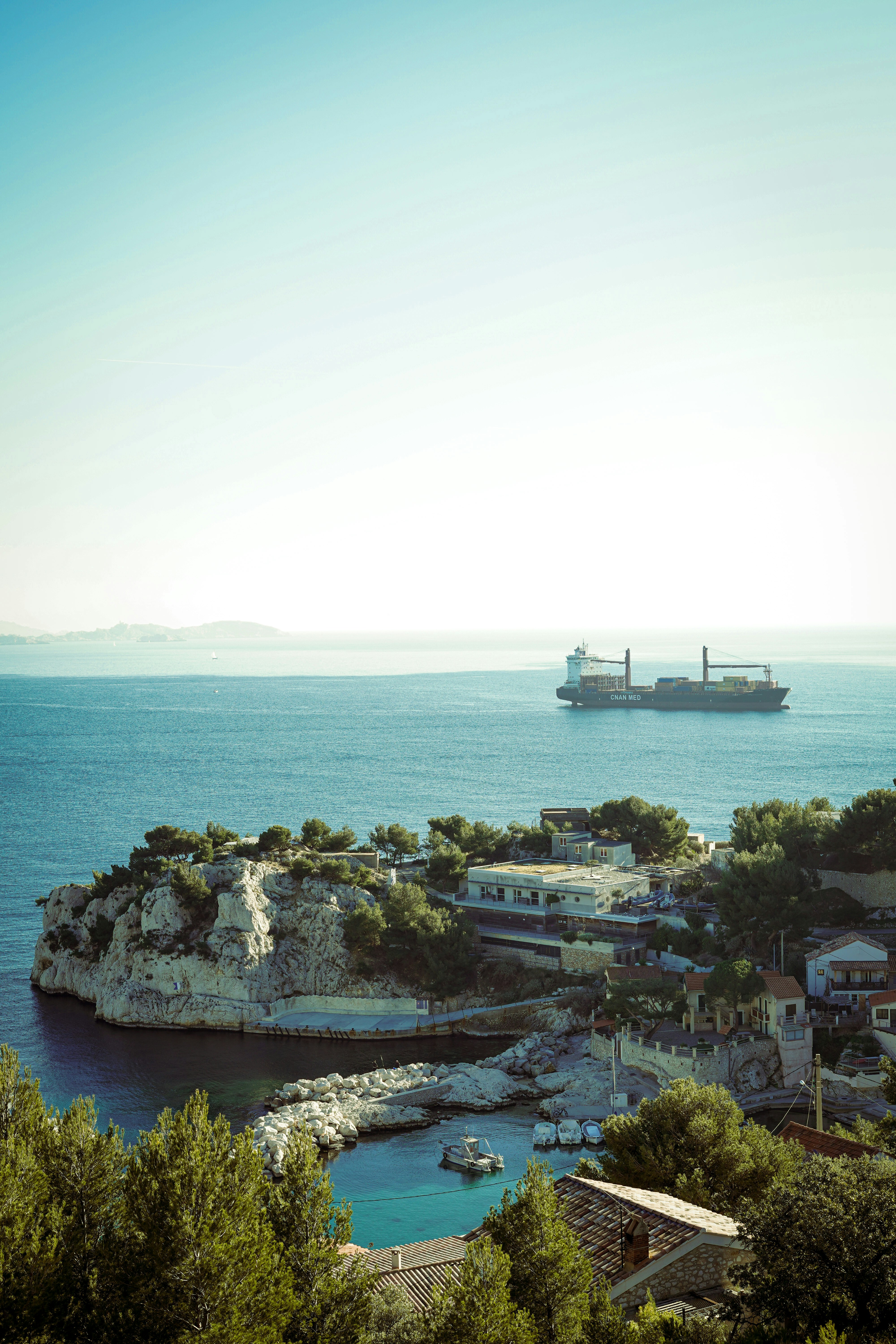 A tranquil coastal scene featuring a cargo ship navigating the calm waters, surrounded by lush greenery and rocky shores.