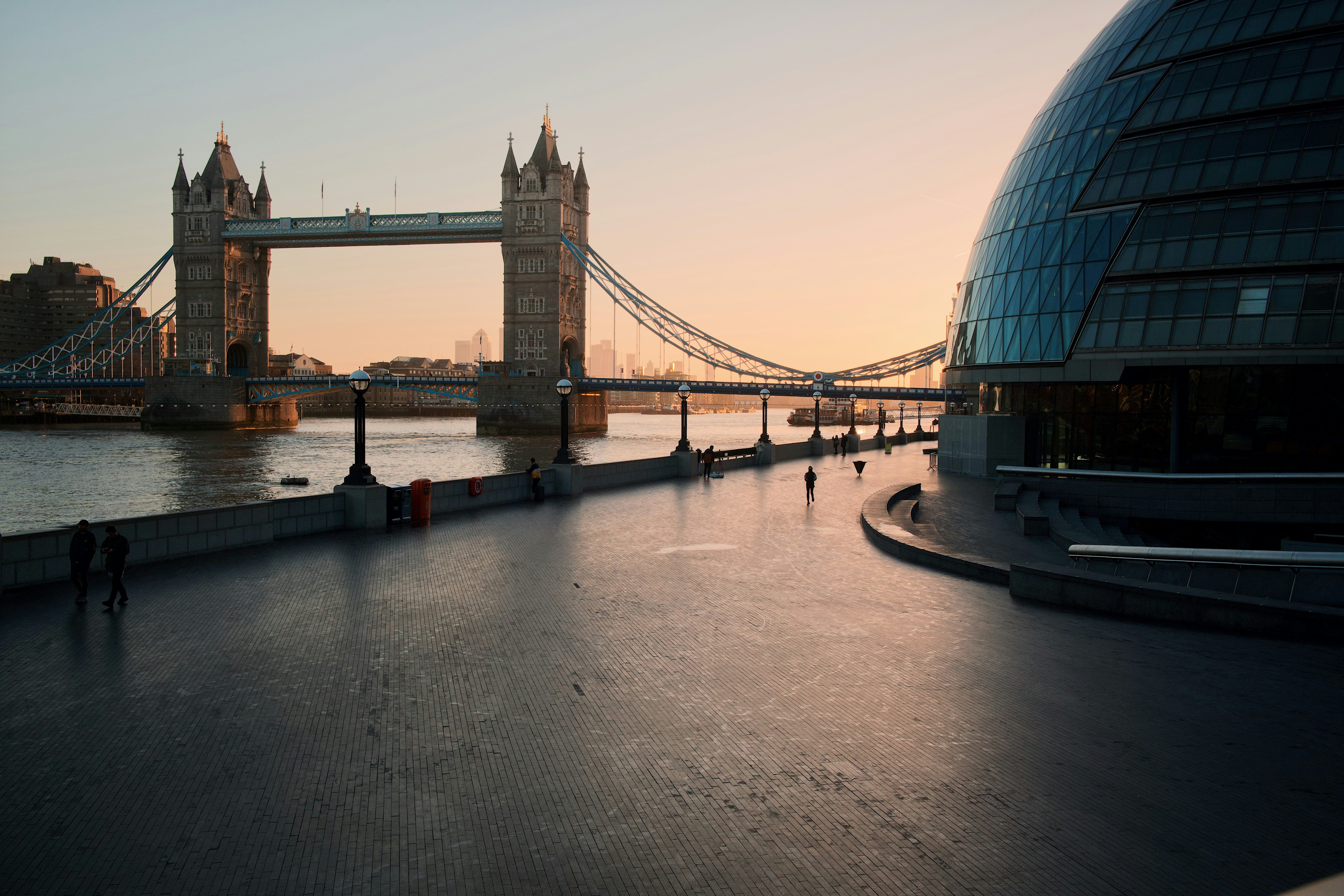 Tower Bridge illuminated at dusk, viewed from the Thames promenade with a modern glass structure in the foreground. The scene captures the blend of historical and contemporary architecture.