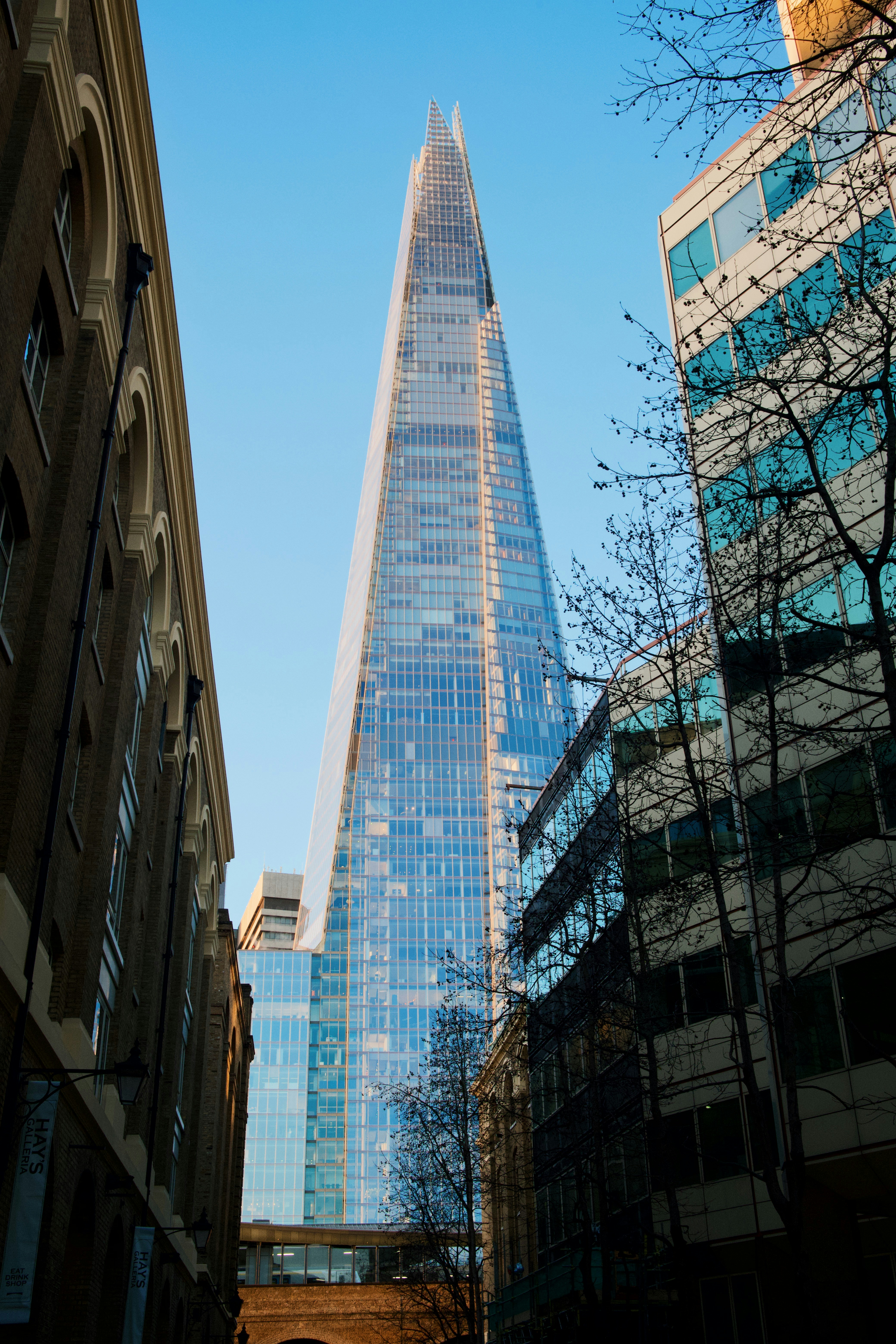 The Shard pierces the sky, framed by surrounding buildings, showcasing its modern architectural design against a clear blue backdrop.
