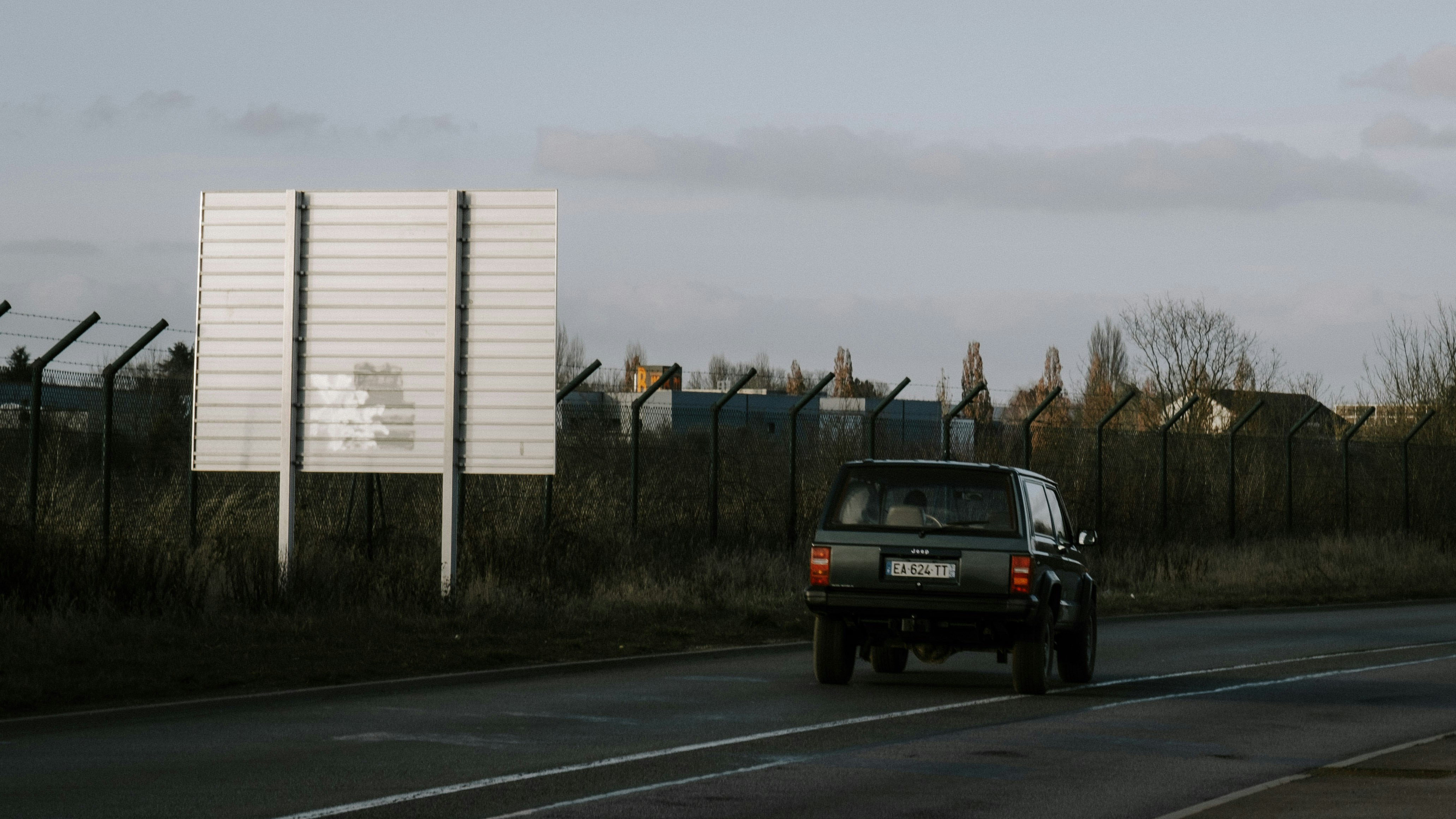 An unmarked roadside sign stands beside a lone vehicle on a quiet street, framed by a chain-link fence and distant industrial structures.