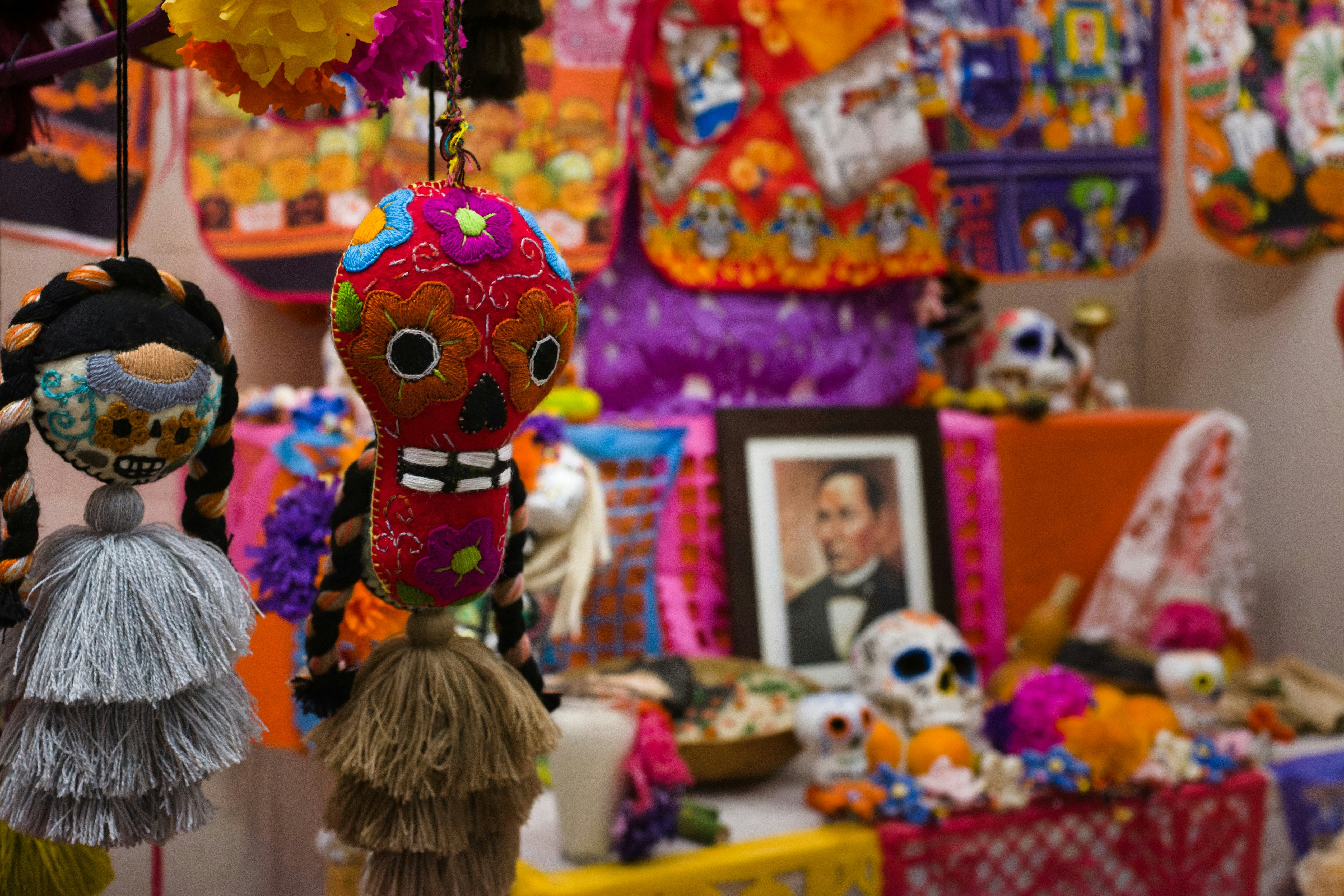 Colorful Skull Ornaments Hanging in an Altar Decorated for Day of the Dead - Two colorful, embroidered skull ornaments hang above a Day of the Dead altar decorated with flowers, candles, skulls, and other traditional decorations. The altar is set up for a celebration of life and remembrance.