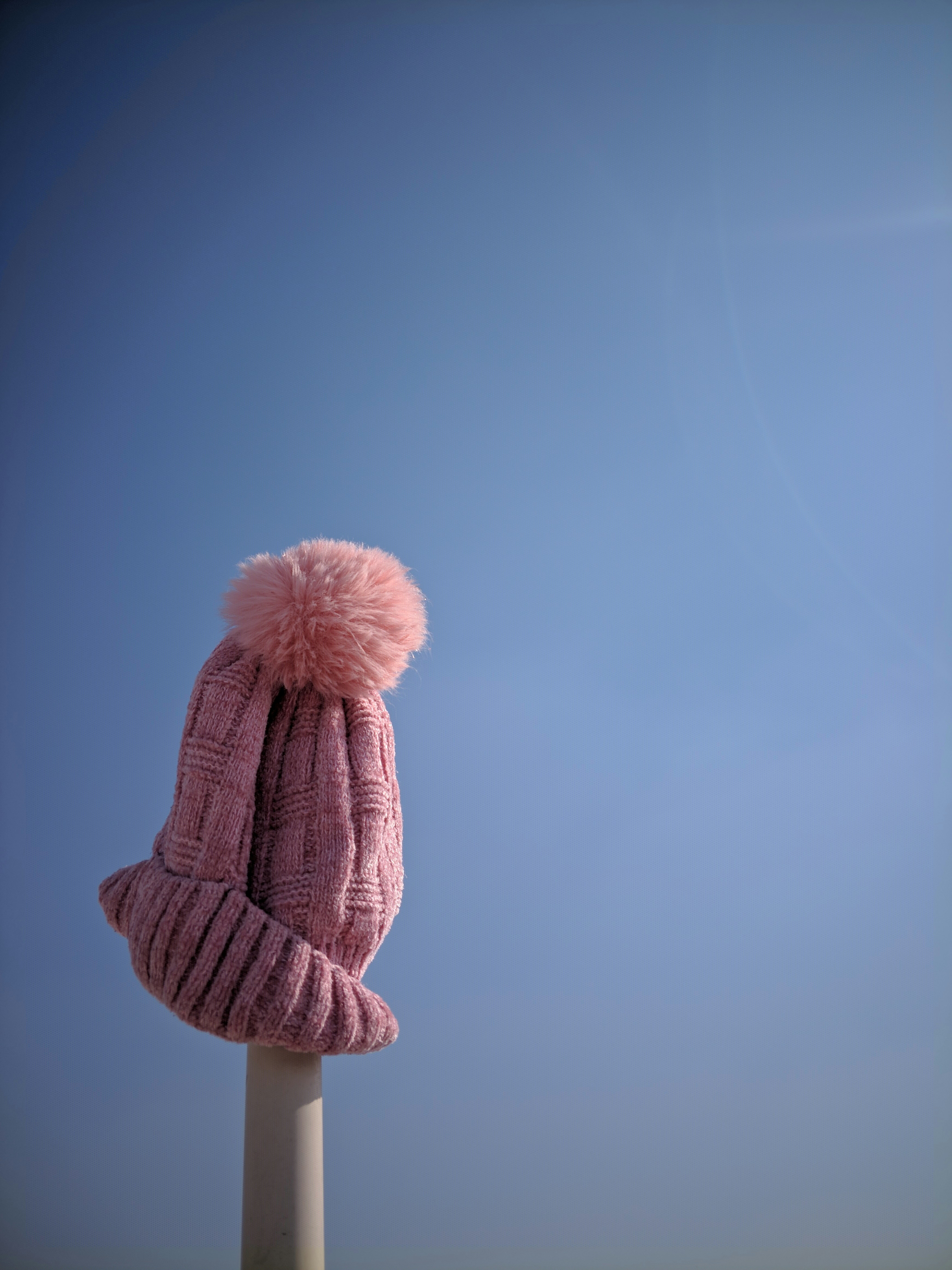 A soft pink knit hat with a fluffy pom-pom perched atop a pole against a clear blue sky.