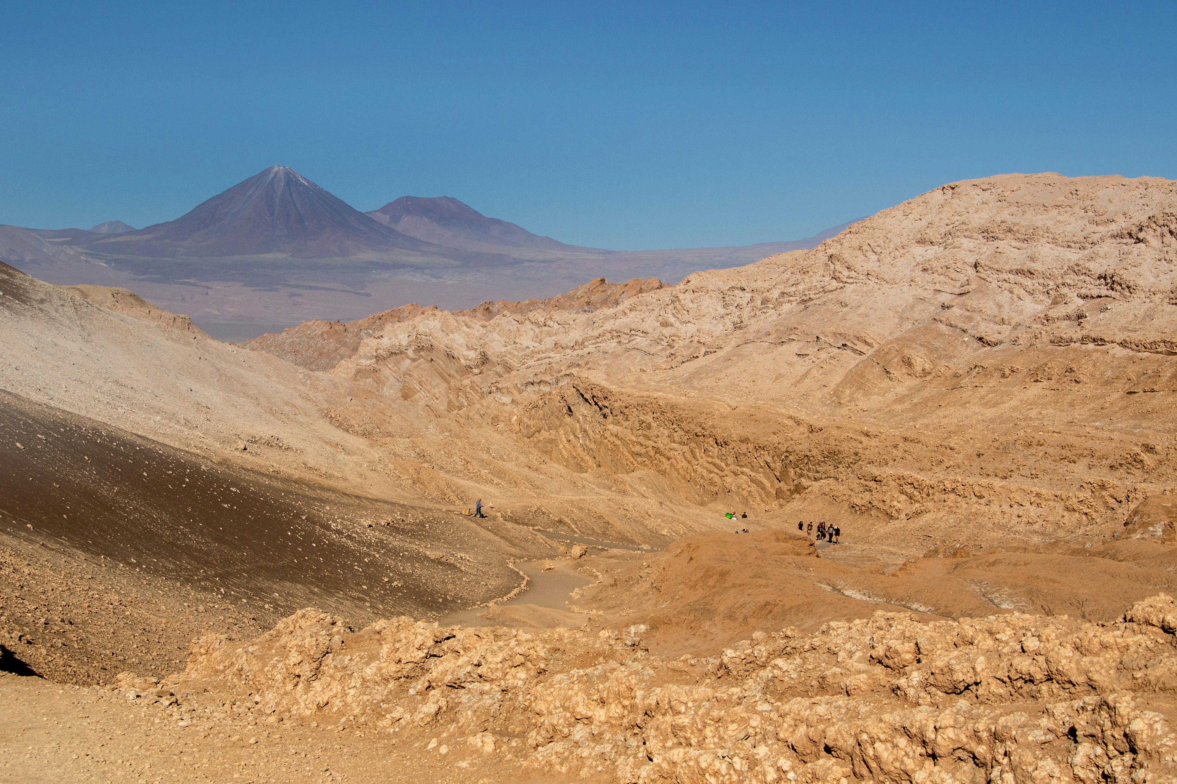 Expansive desert landscape with rugged mountains in the background, showcasing hikers traversing the arid terrain under a clear blue sky.