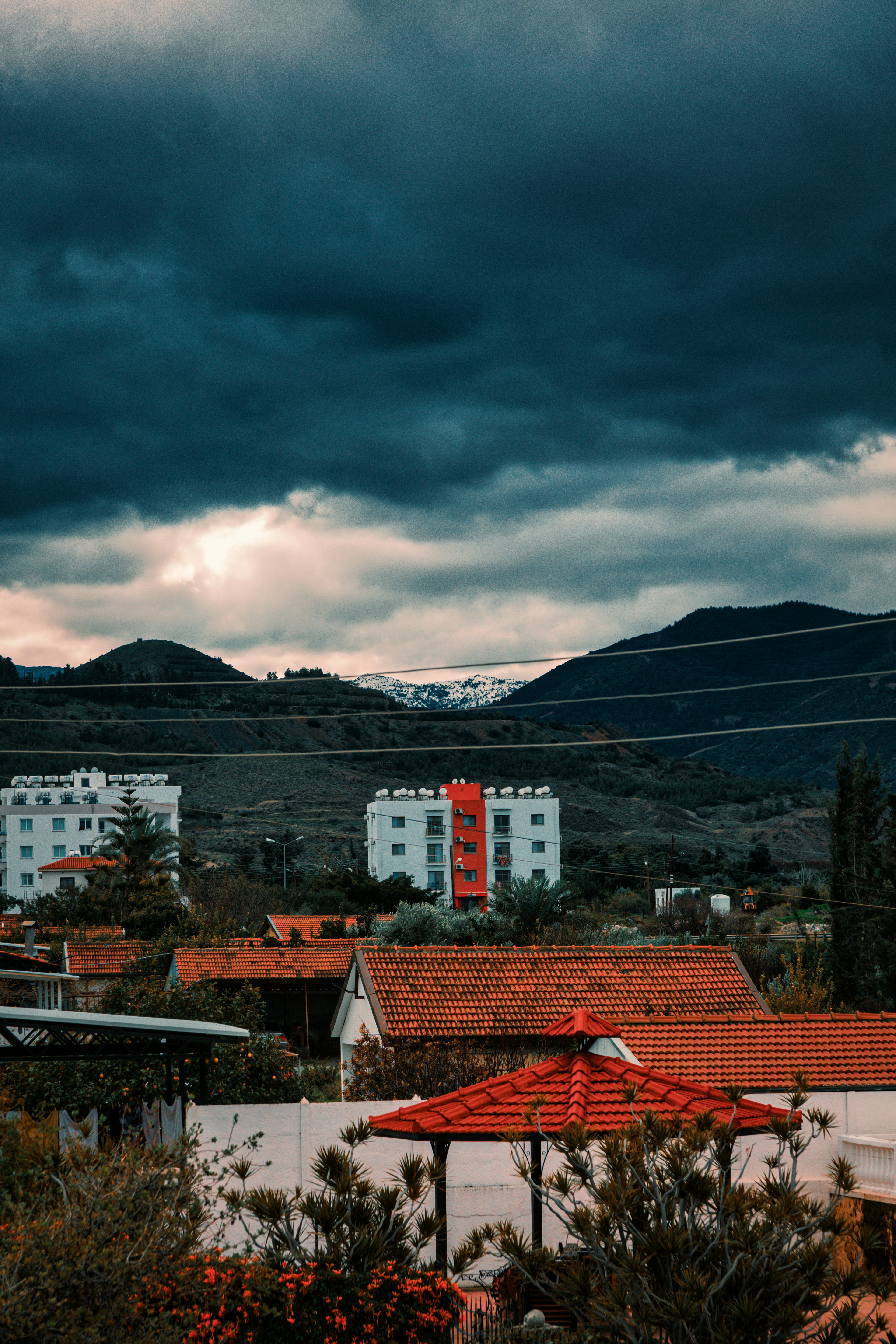 A vibrant red building stands out amidst a landscape of rooftops and distant mountains under a moody sky. Snow-capped peaks loom in the background.