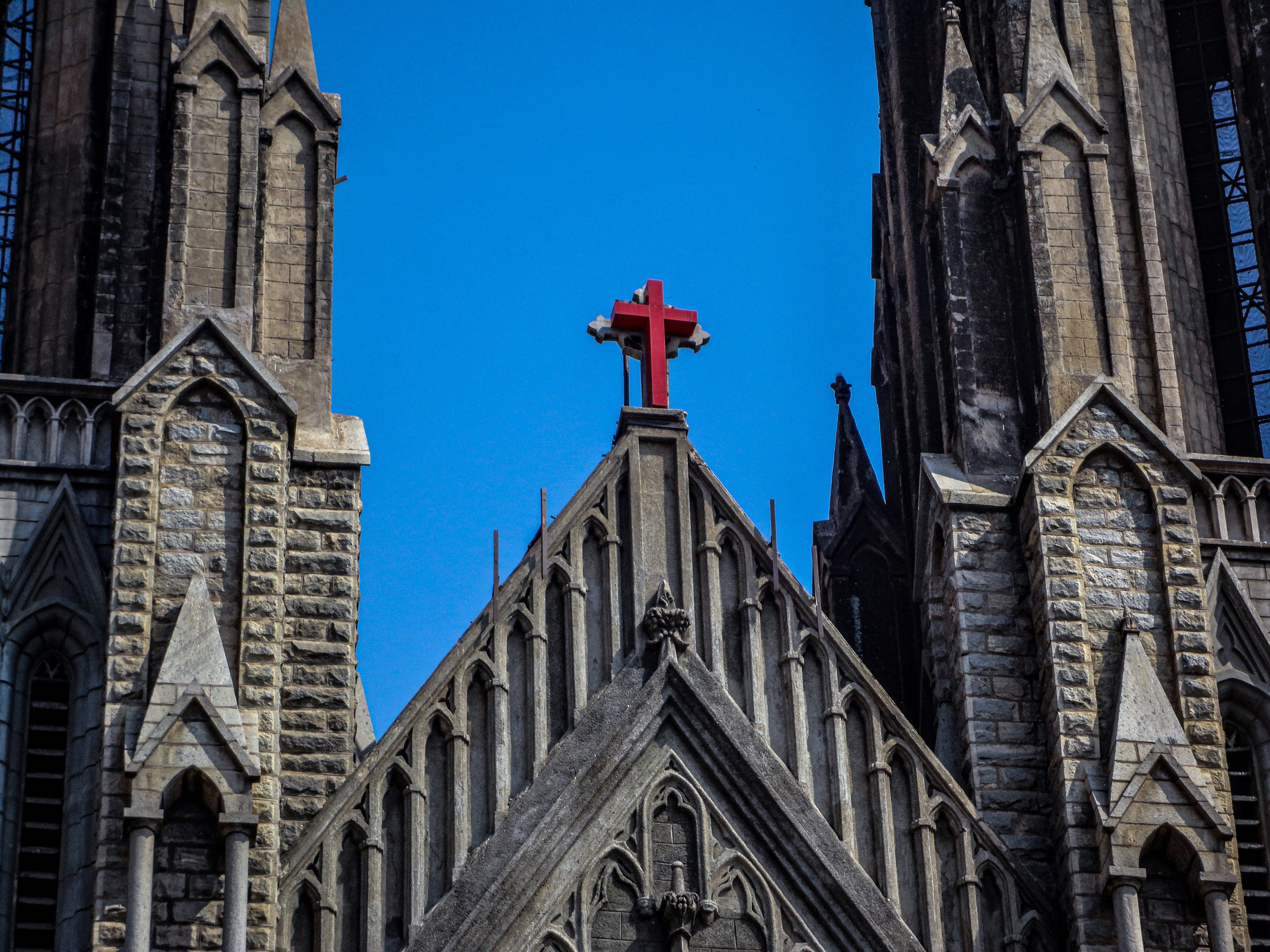 Photograph of a Gothic cathedral's central gable with a red cross atop carved stone, set against a bright blue sky.