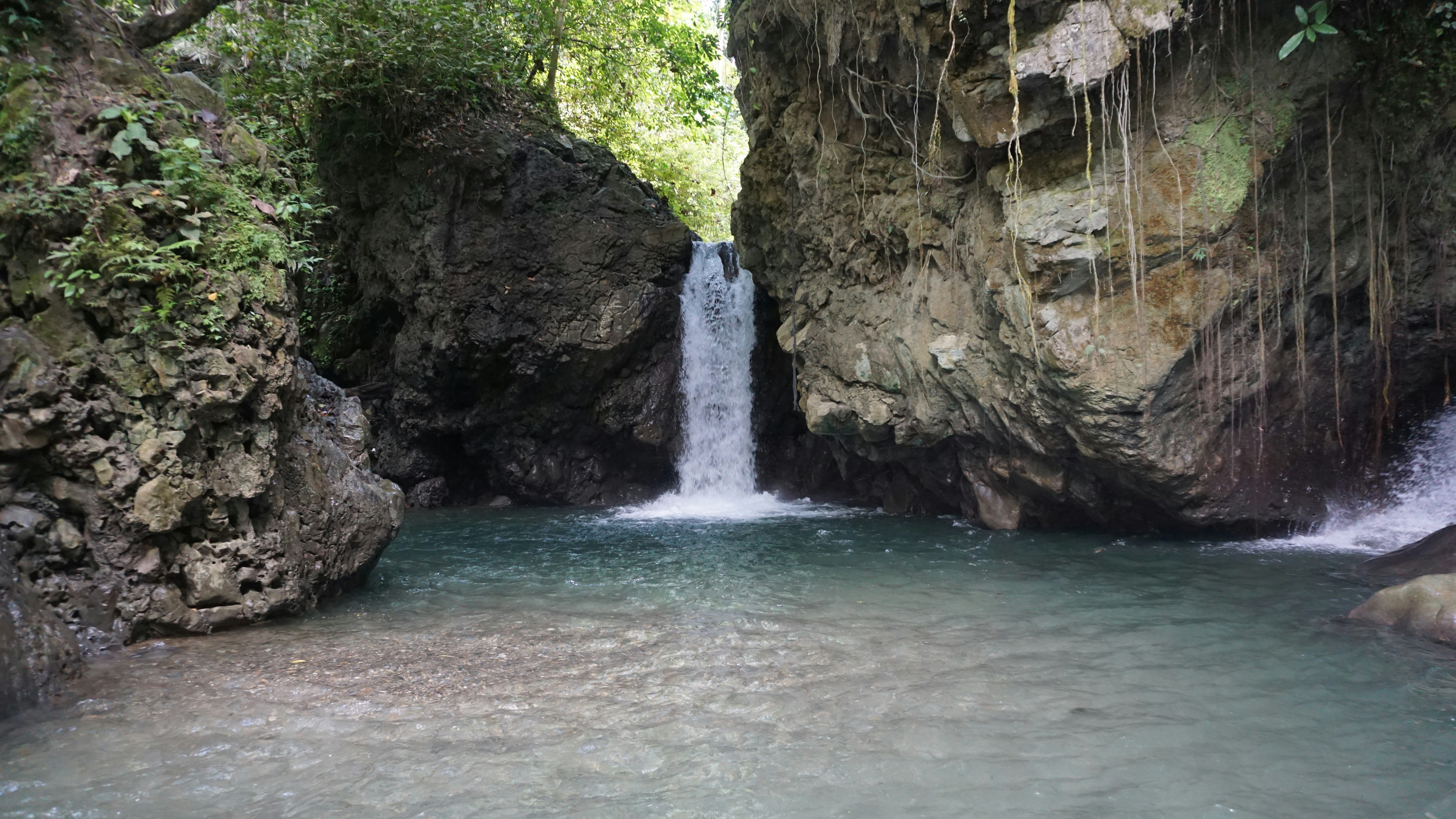 A serene waterfall cascading between rugged rock formations, surrounded by lush greenery and clear blue water.