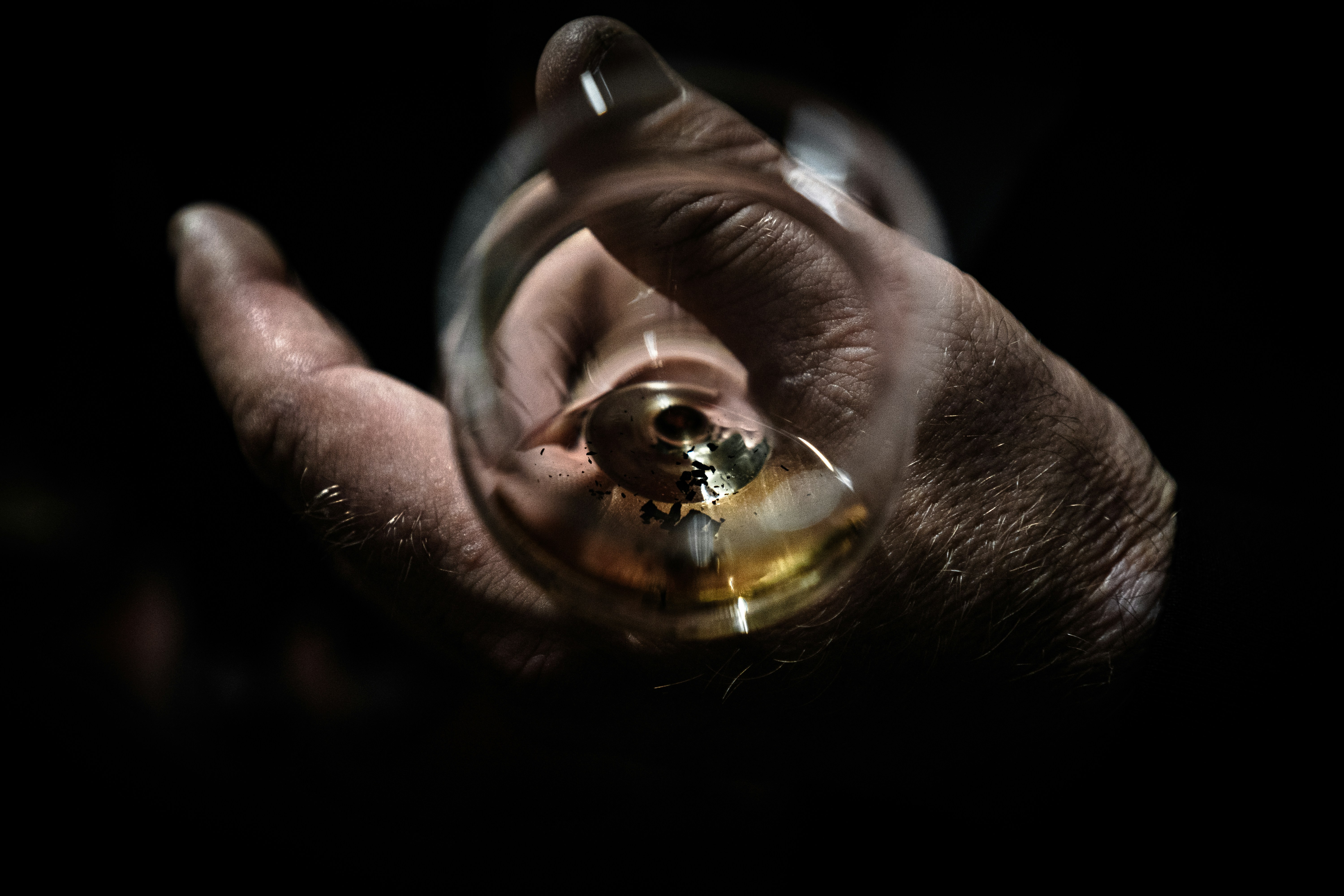 A hand cradling a glass of amber liquid, capturing reflections and depth against a dark background.
