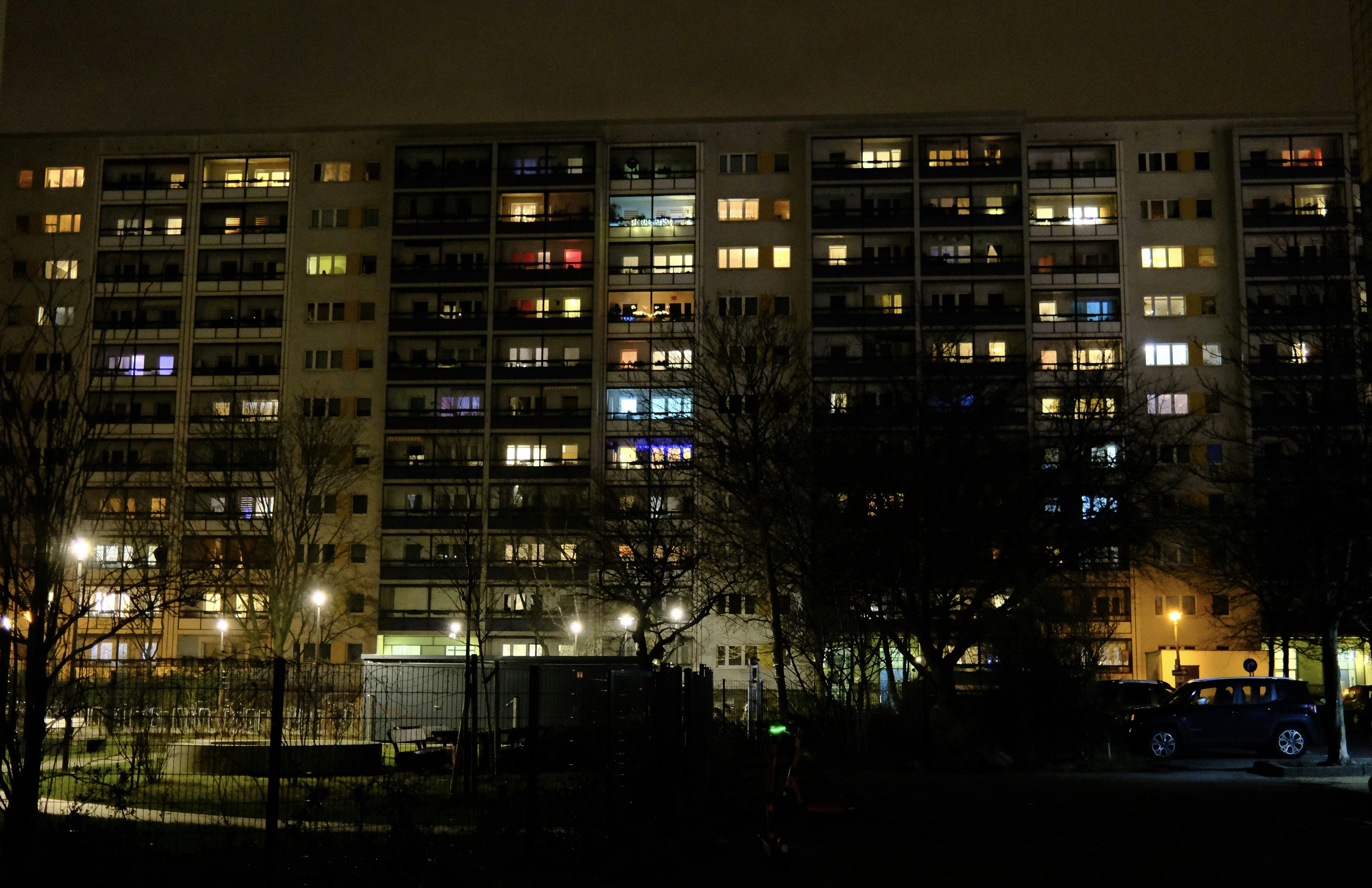 Illuminated apartment building at night, showcasing various lit windows and silhouettes of residents. The scene captures the essence of urban living after dark.