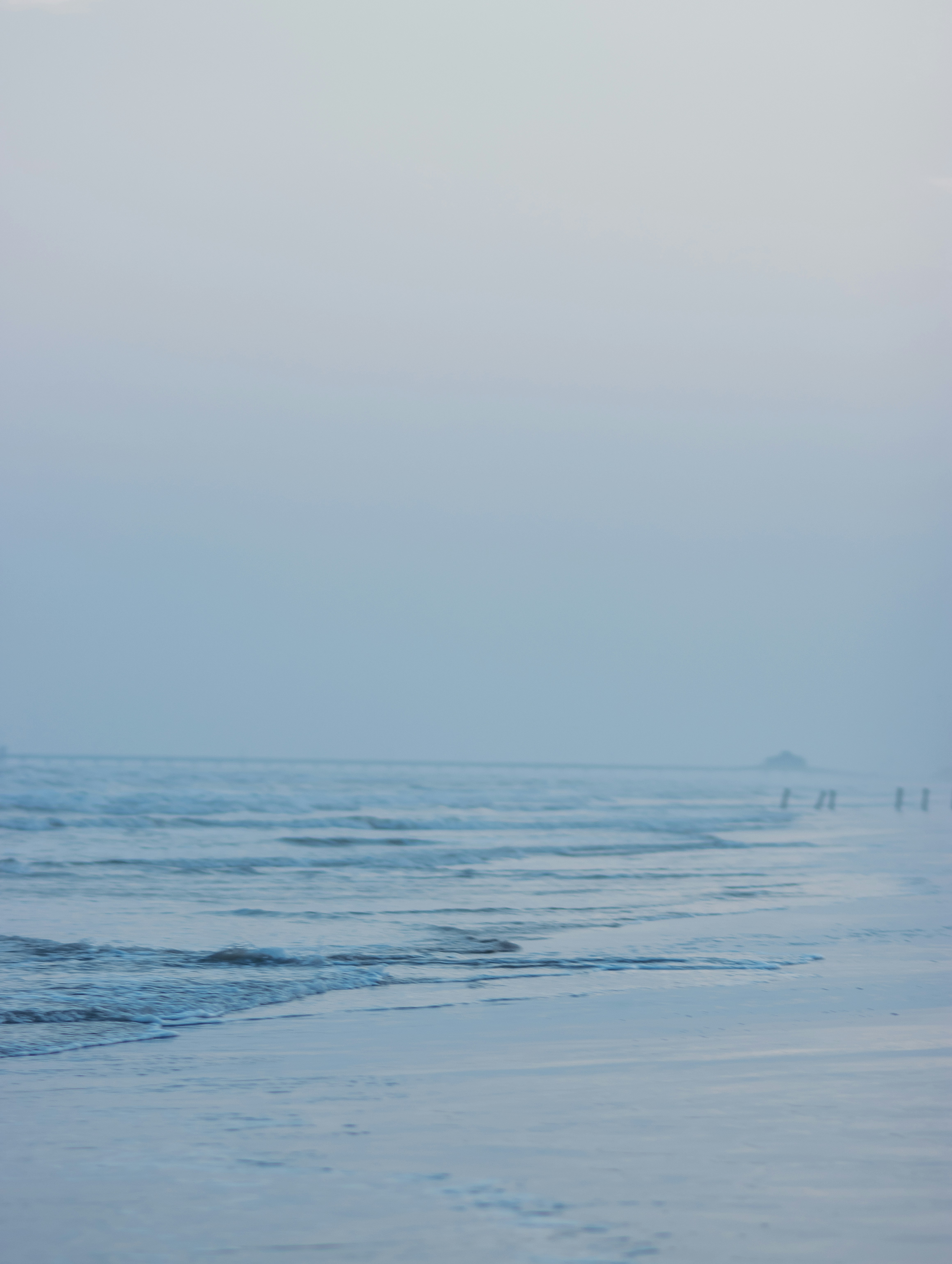 Gentle waves caress the shore under a soft, hazy sky, creating a tranquil seascape at dusk.
