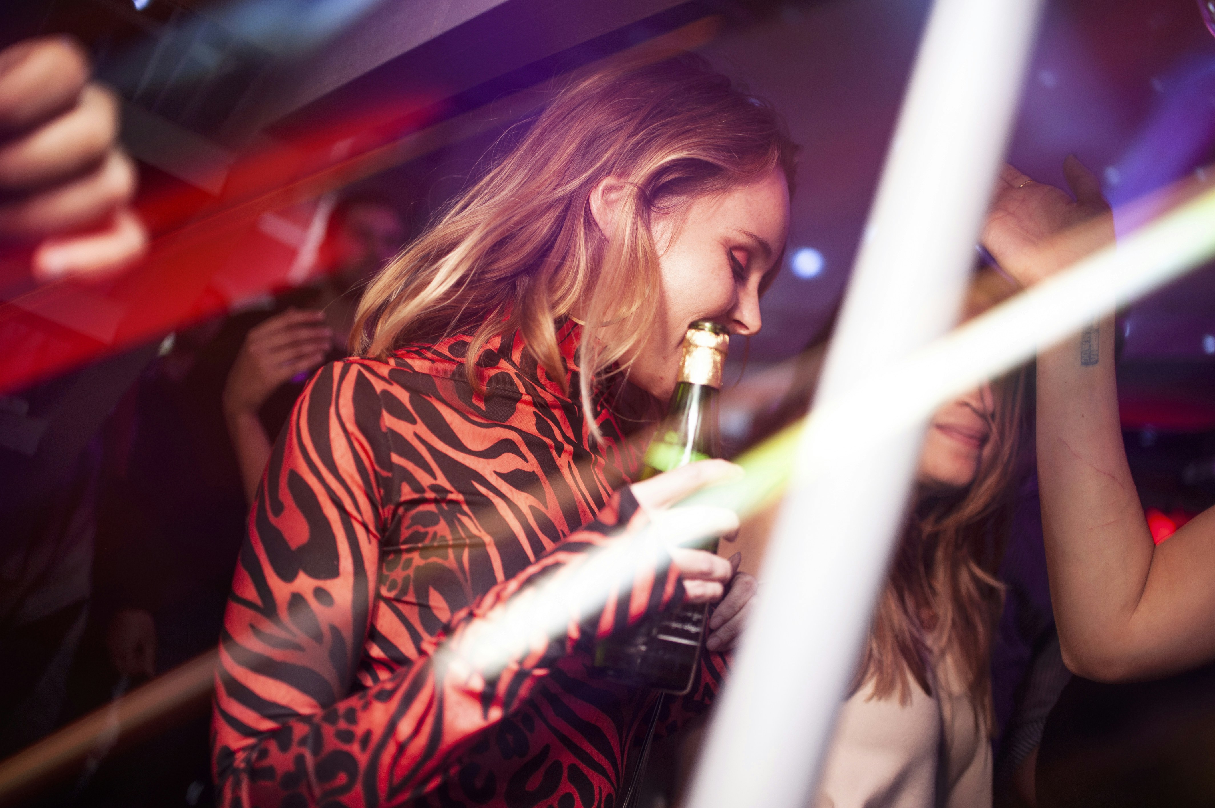 Two women enjoying a vibrant night out, one holding a drink while the other dances. The atmosphere is enhanced by colorful lights and a sense of camaraderie.