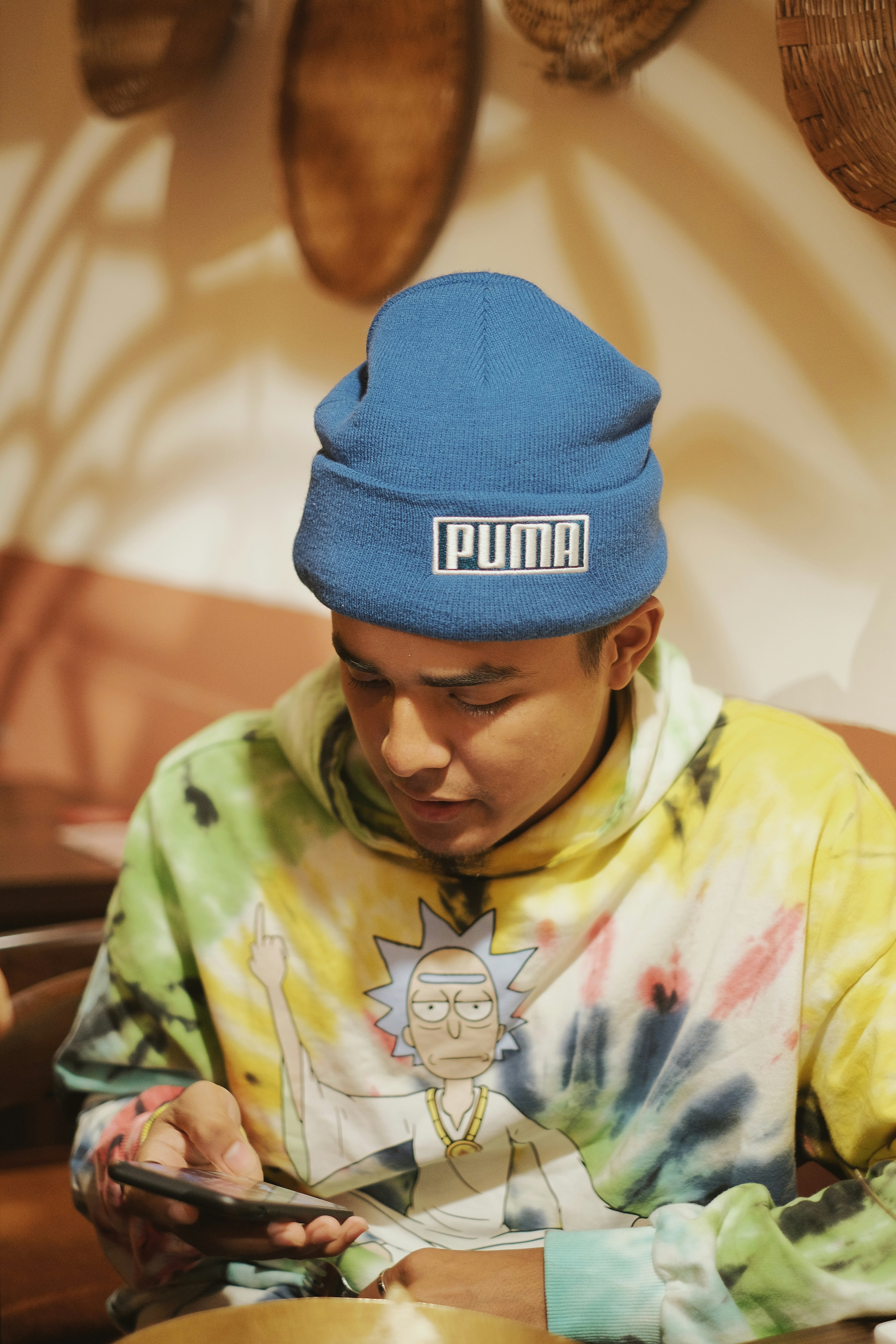 Portrait of a person in a blue Puma beanie scrolling a smartphone, wearing a tie-dye hoodie with a Rick and Morty cartoon graphic.