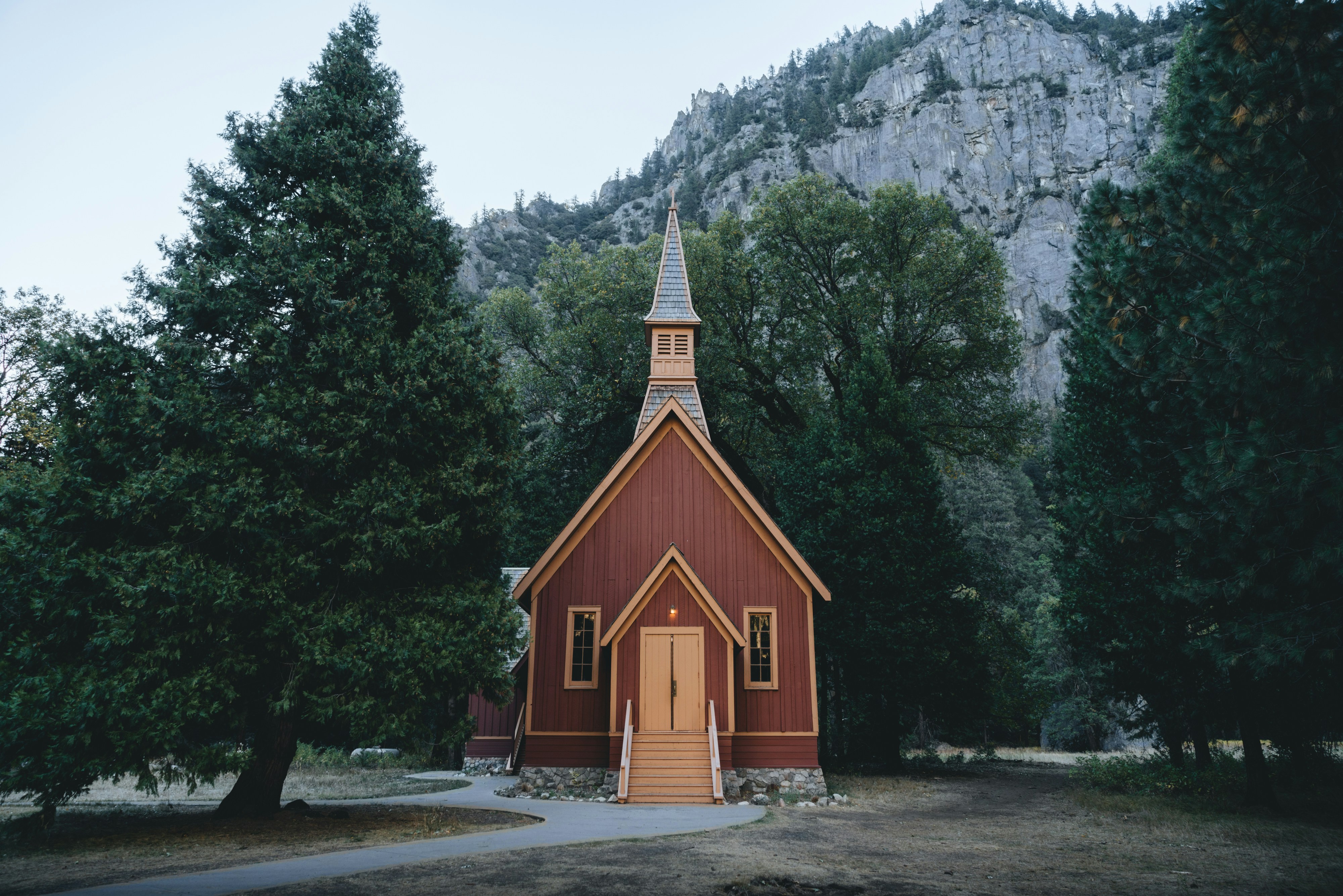 Charming red chapel nestled among towering trees with a rocky mountain backdrop, showcasing a serene atmosphere.