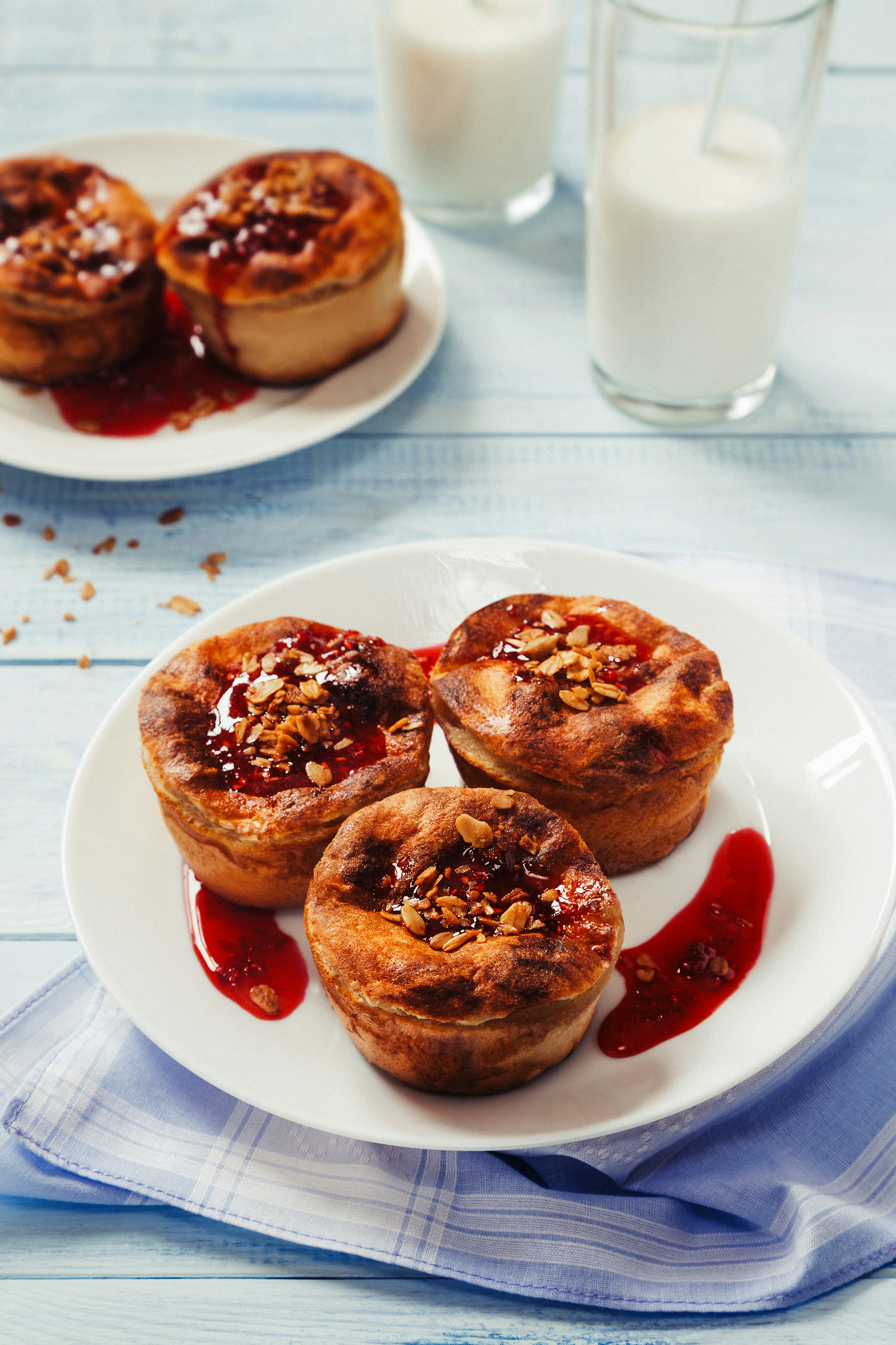 Three dessert cups filled with fruit compote and topped with granola, served on a white plate with a drizzle of sauce. A refreshing glass of milk is in the background.