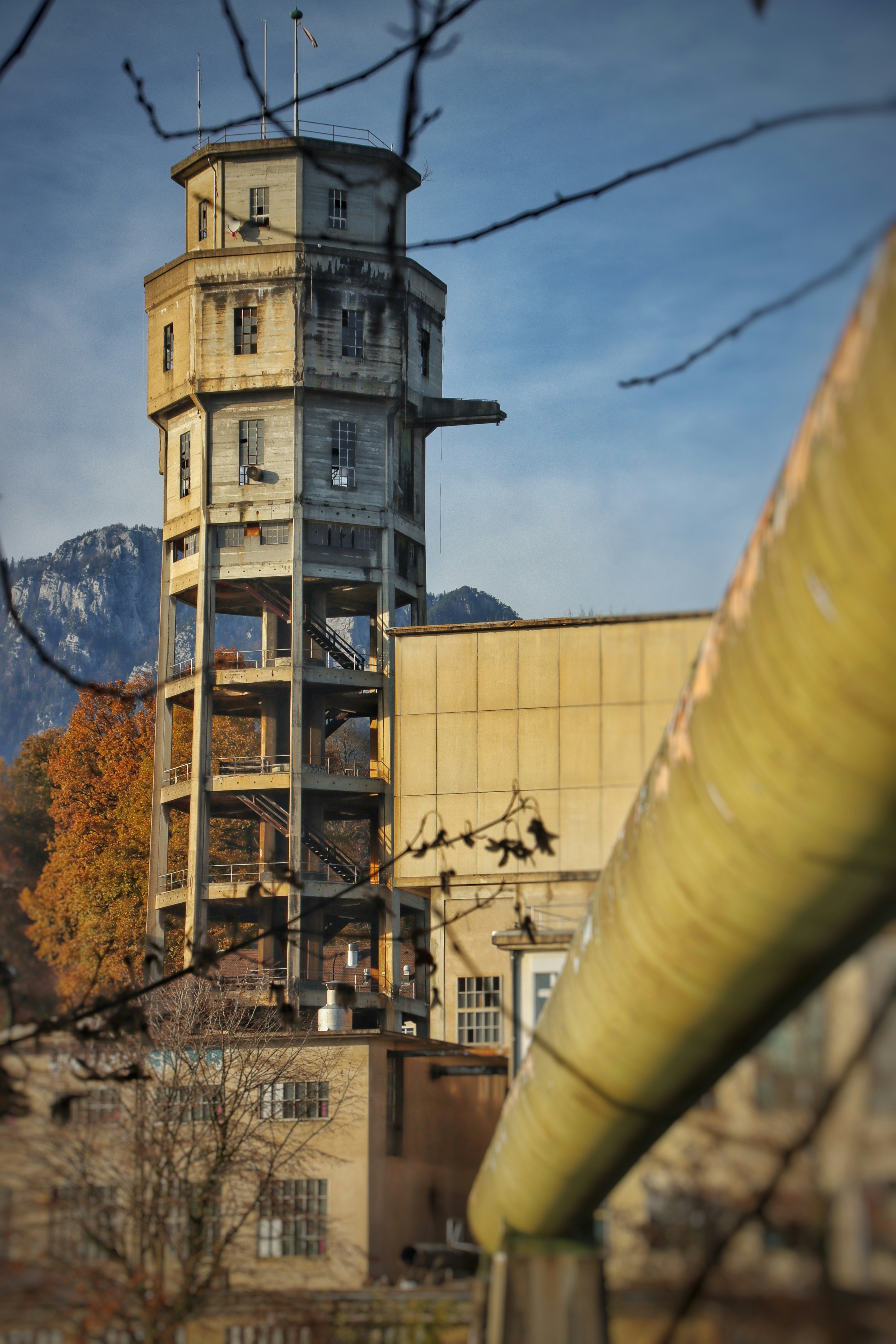 Abandoned industrial tower surrounded by autumn foliage, with a pipeline leading into the frame. The backdrop features distant mountains under a clear sky.
