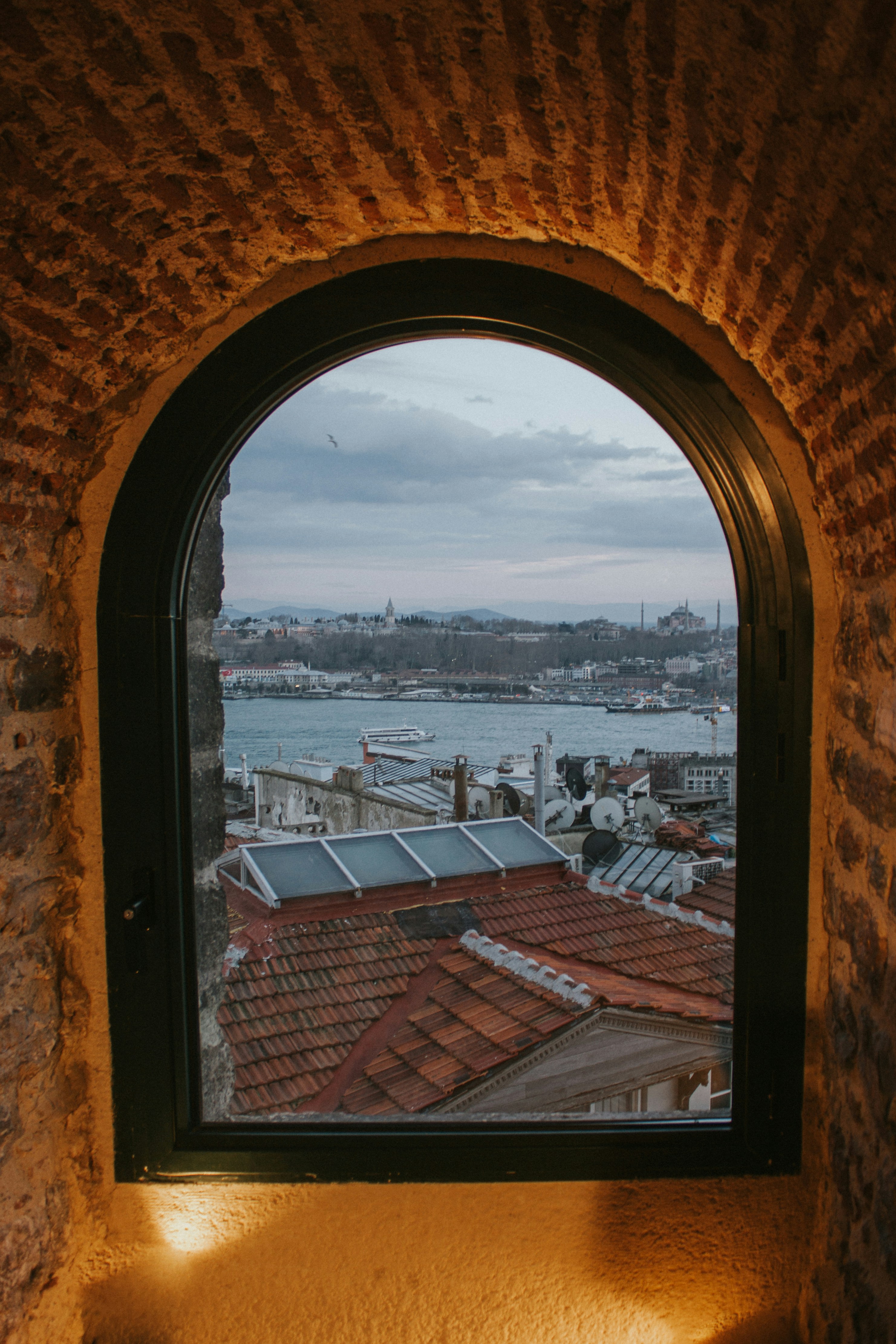 View from a stone-framed window showcasing rooftops and the serene waters of Istanbul, with distant hills under a cloudy sky.