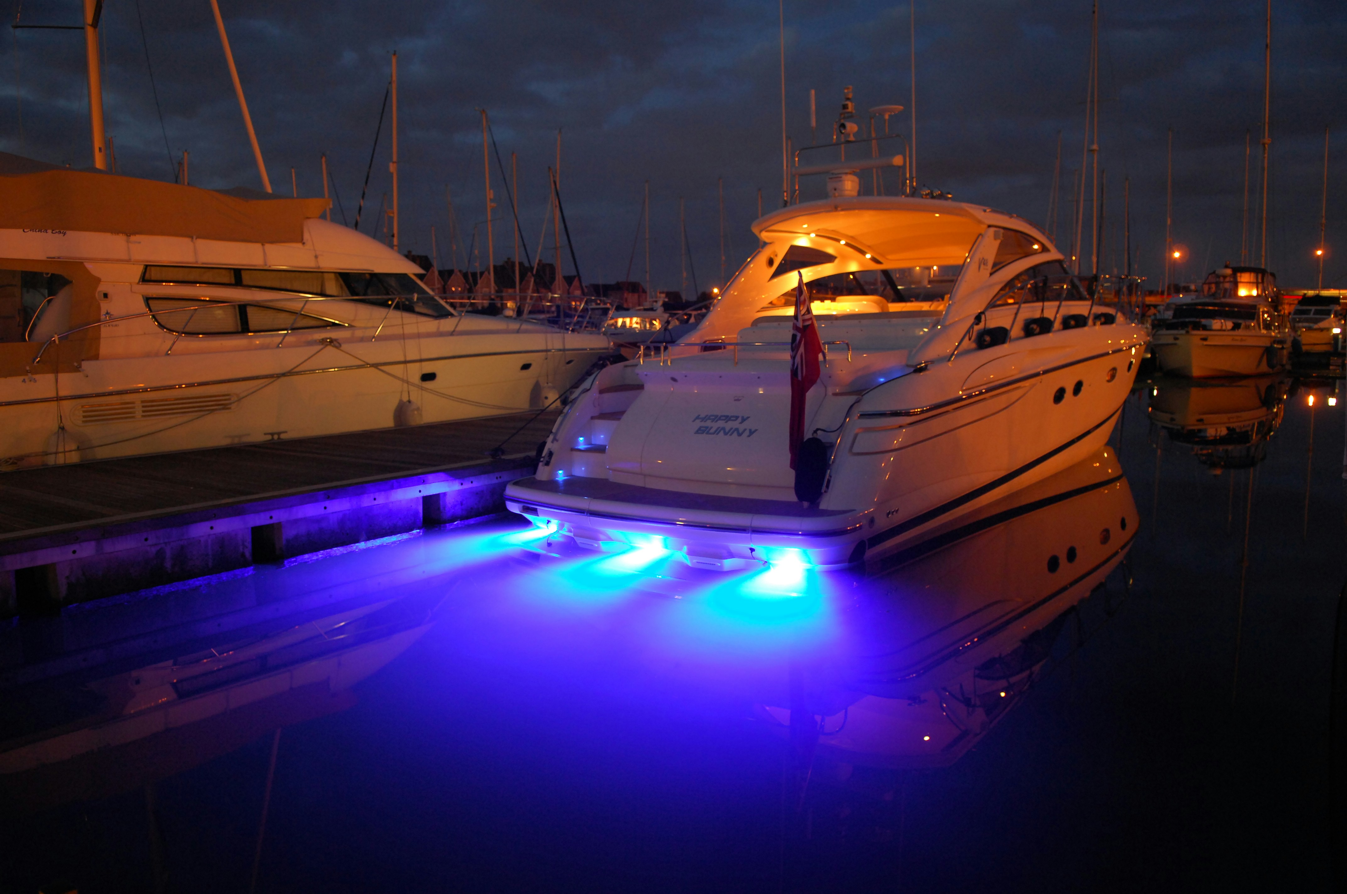 Luxury boat anchored at a marina, illuminated by vibrant blue lights reflecting in the calm water. The scene captures a tranquil evening atmosphere.